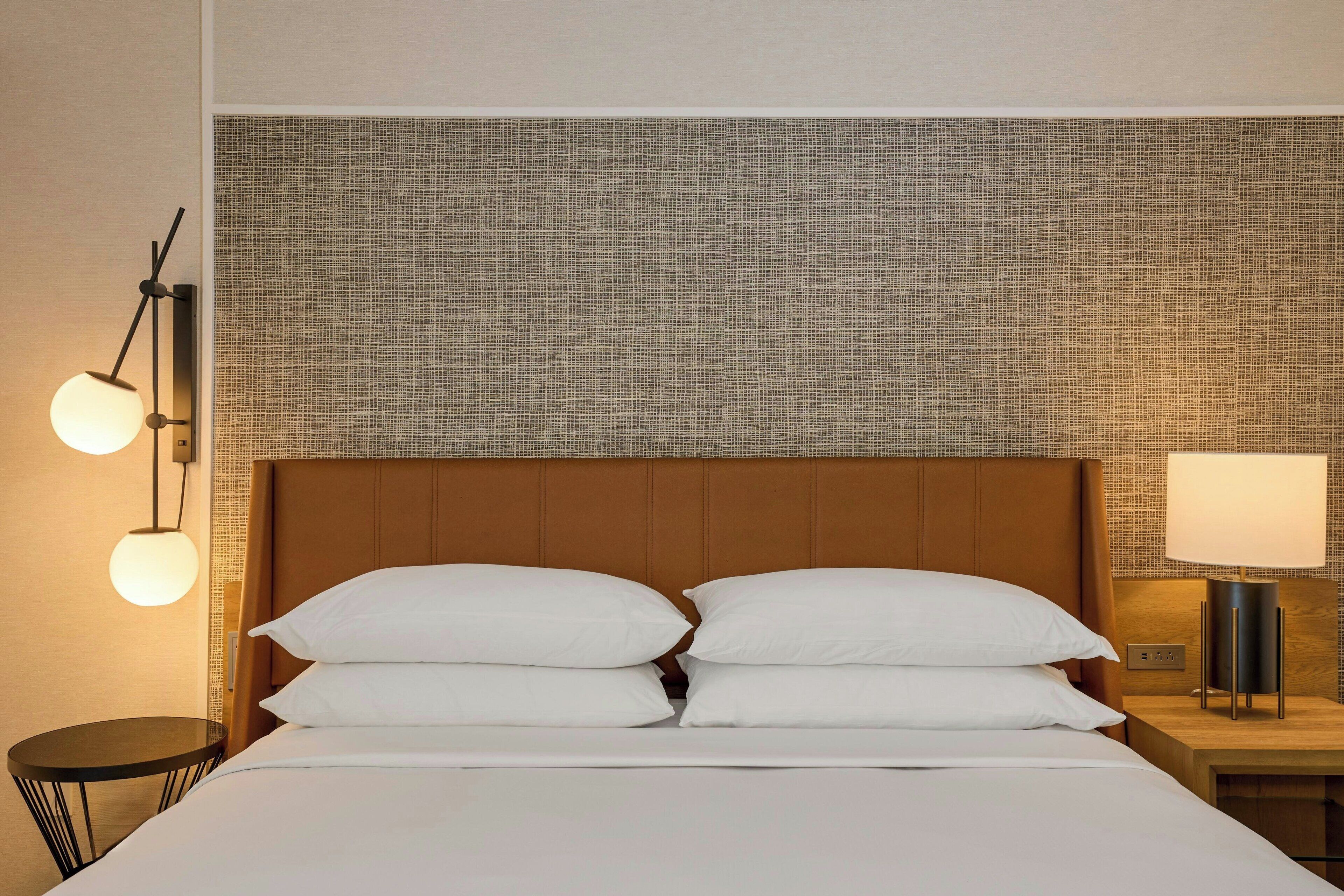 Quarto de hotel com uma cama grande, duas luminárias de cabeceira, uma mesa de cabeceira e um painel de parede texturizado.