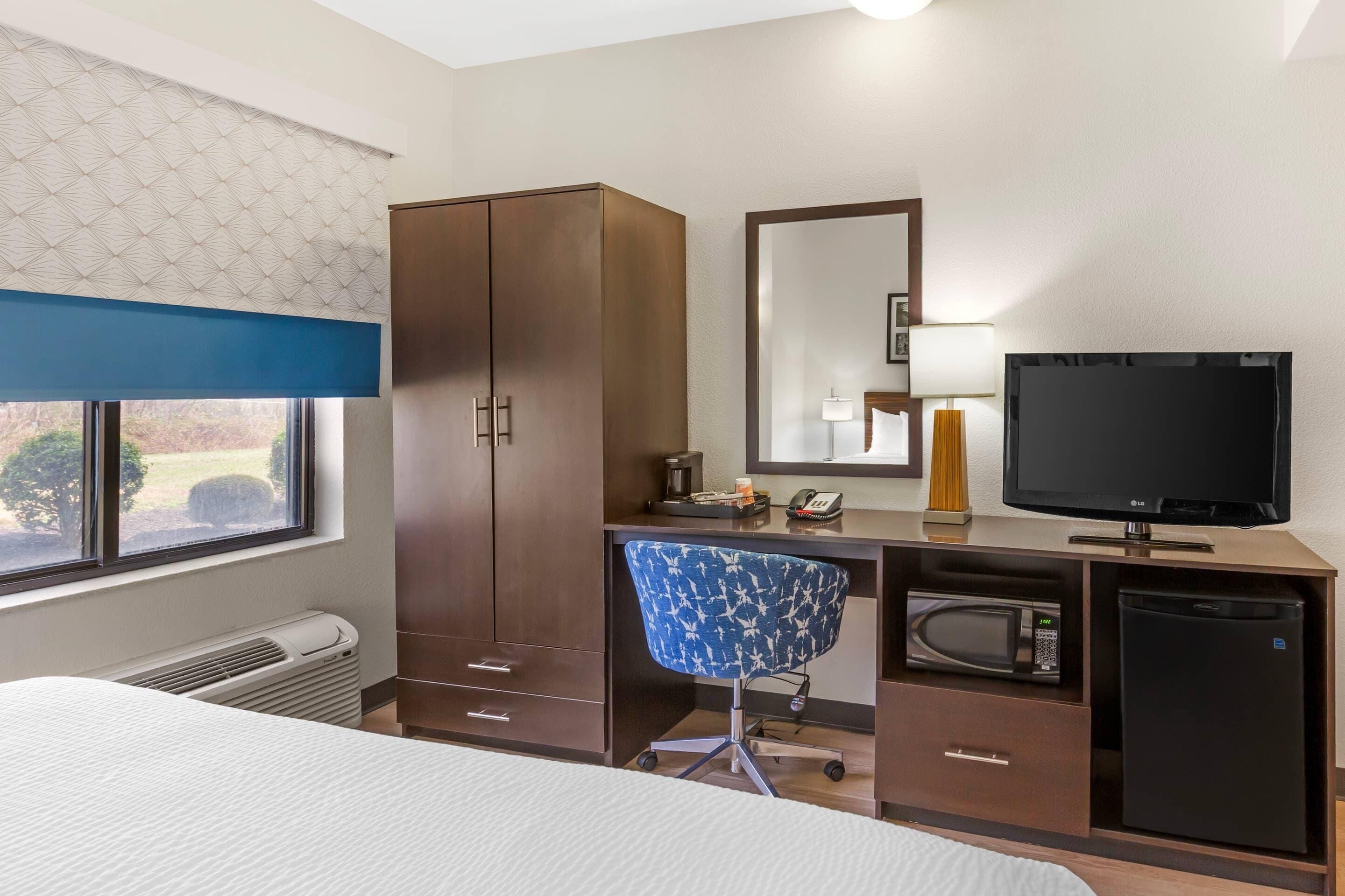 Quarto de hotel com cama, escrivaninha com computador, televisão, micro-ondas e frigorífico.