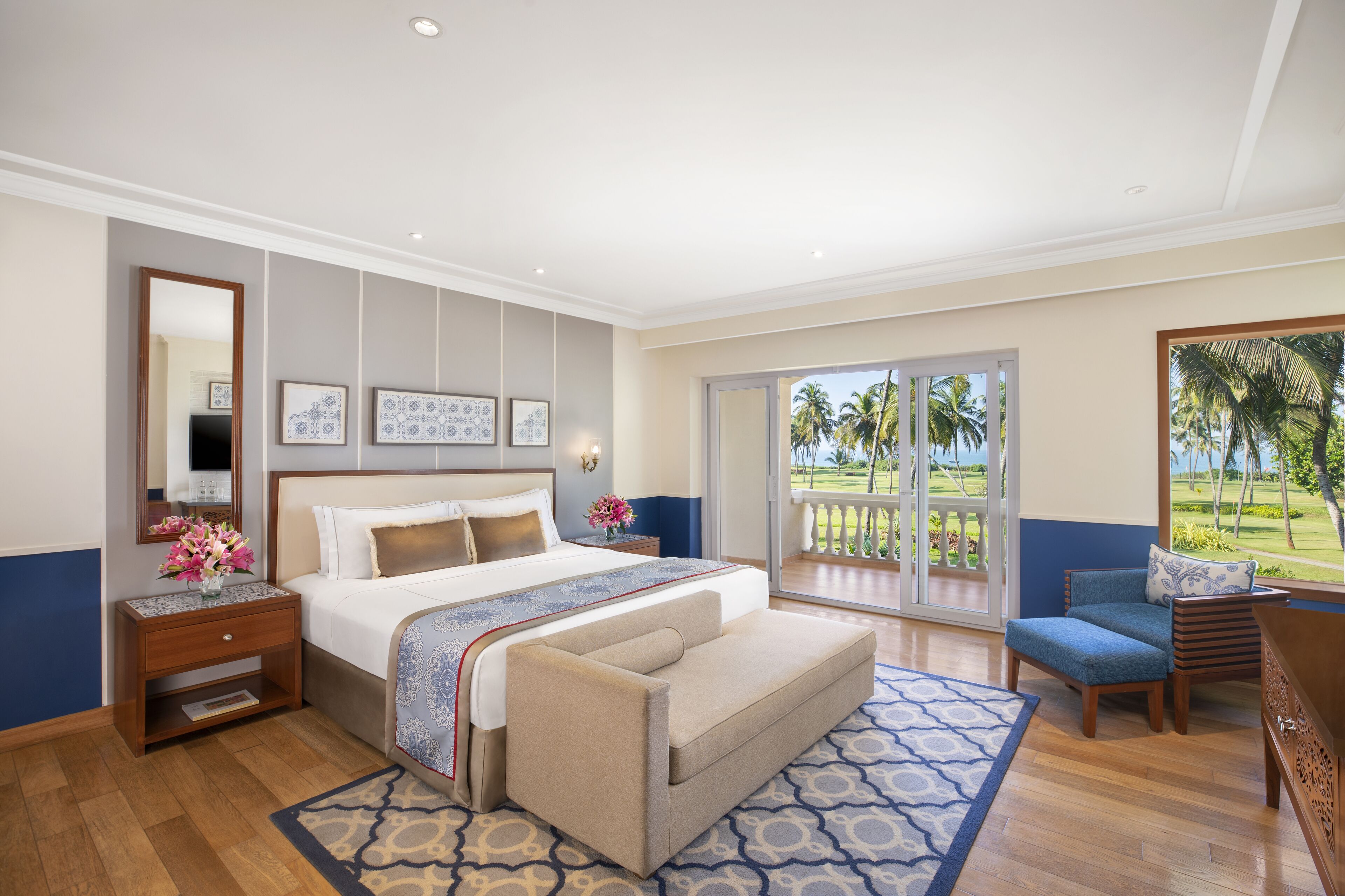 Quarto de hotel com uma cama grande, área de estar com sofá e poltrona, mesa de cabeceira de madeira, espelho e vista para palmeiras através de uma porta de correr de vidro.