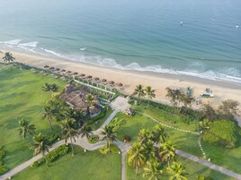 Vista aérea de um resort à beira-mar, com uma construção de telhado de palha, palmeiras e um caminho que leva à praia.