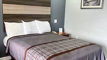 Cama bem arrumada com colcha estampada e almofadas, cabeceira de madeira, mesa de cabeceira com controle remoto e um quadro na parede.