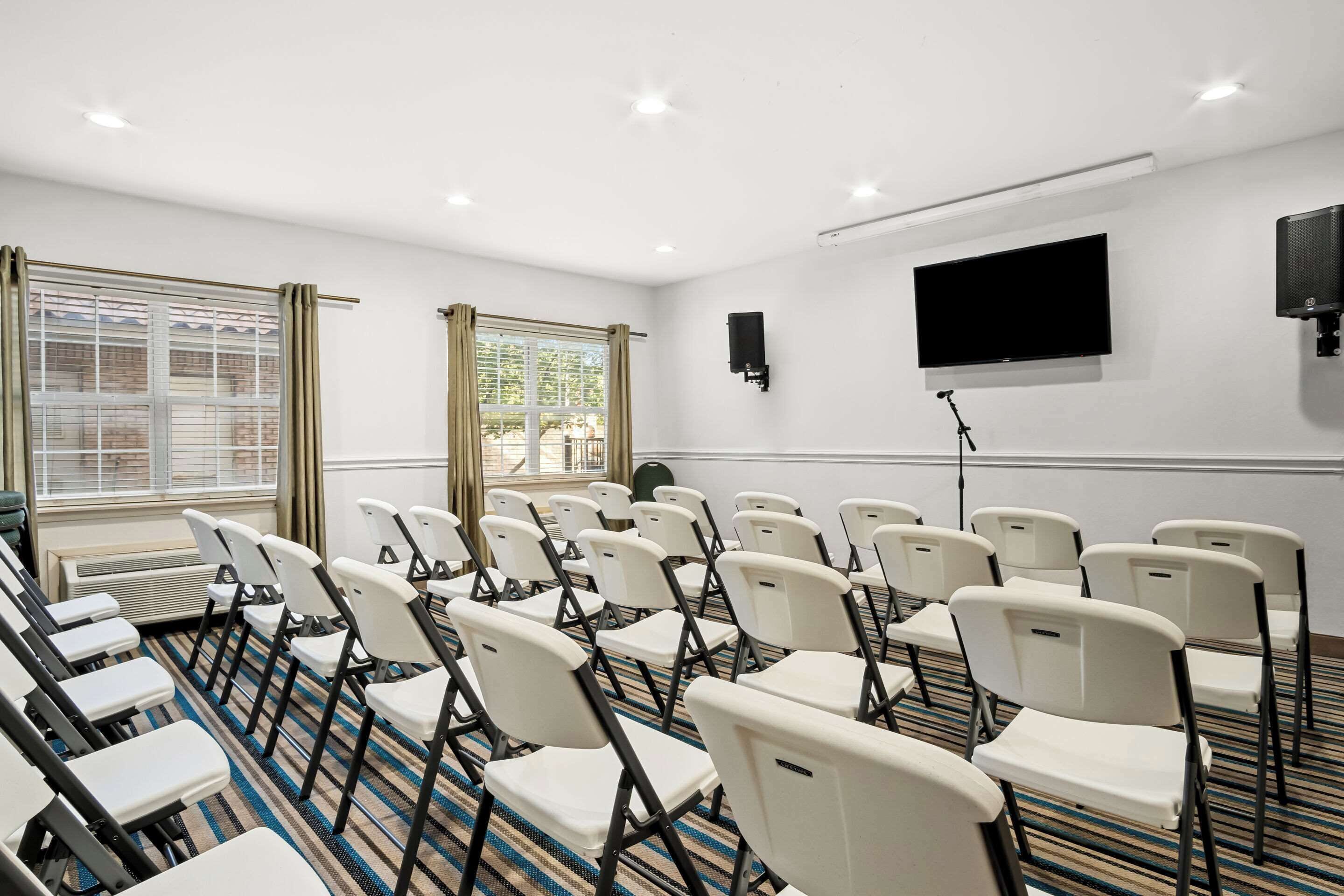 Uma sala de conferências com fileiras de cadeiras brancas, uma televisão de tela plana grande fixada na parede e uma janela com cortinas.