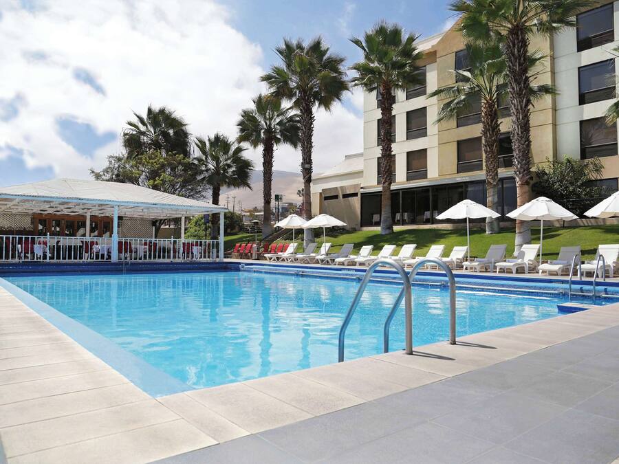 Piscina com espreguiçadeiras e guarda-sóis, cercada por palmeiras e um edifício com vários andares.