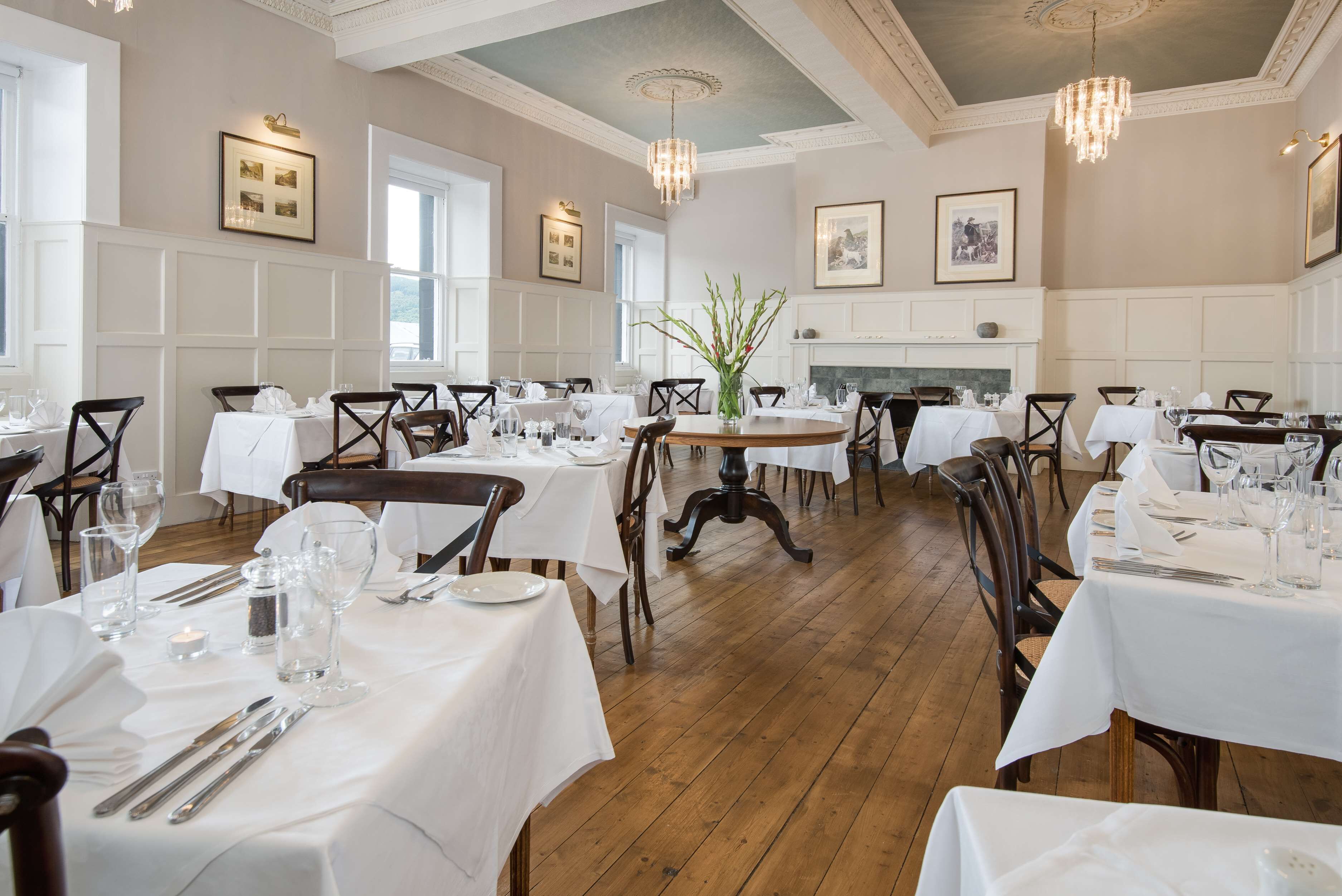 Uma sala de jantar com várias mesas preparadas para o serviço, piso de madeira e lustres.