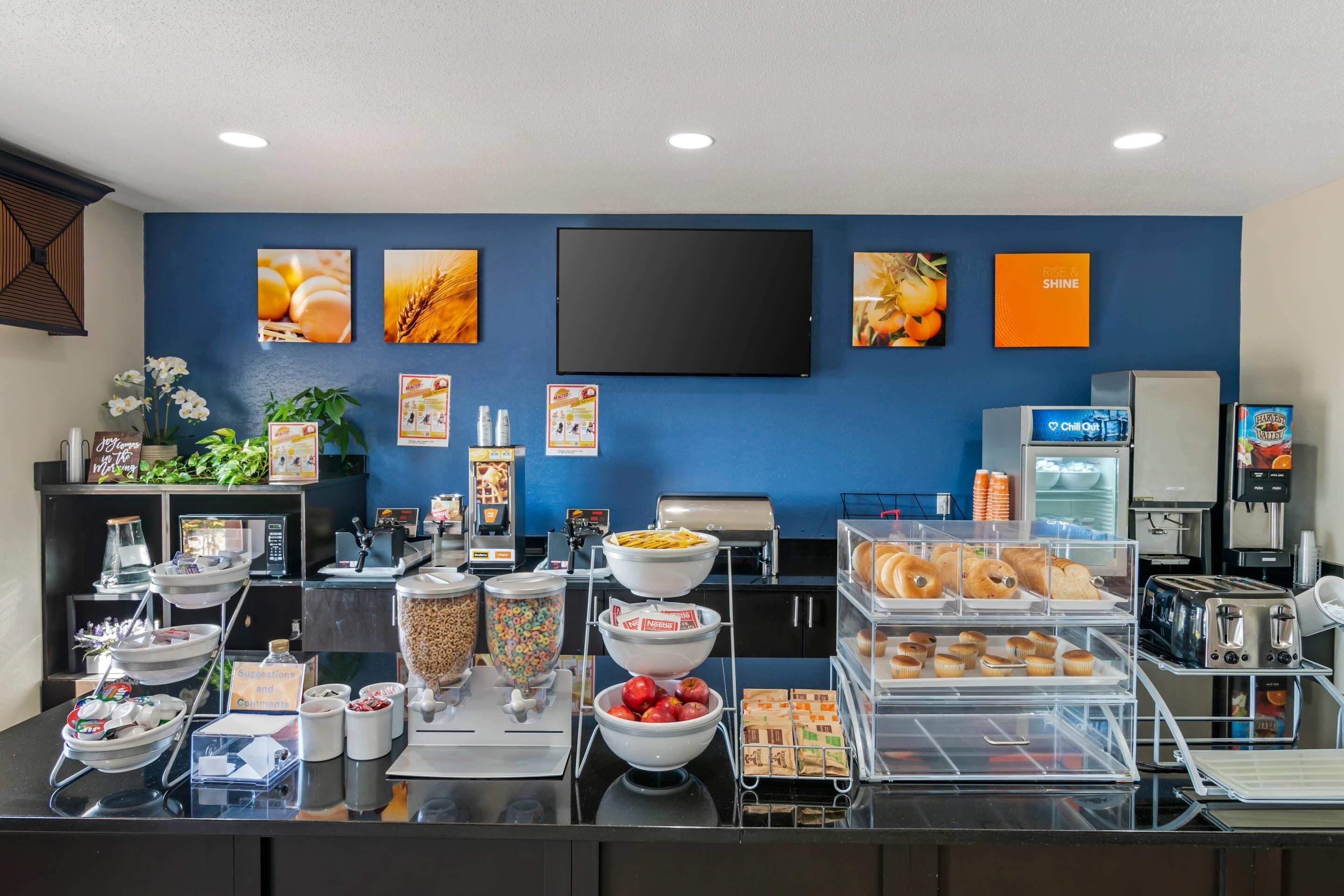 Buffet de café da manhã com diversas opções de comida, um espaço para café e uma TV fixada na parede.