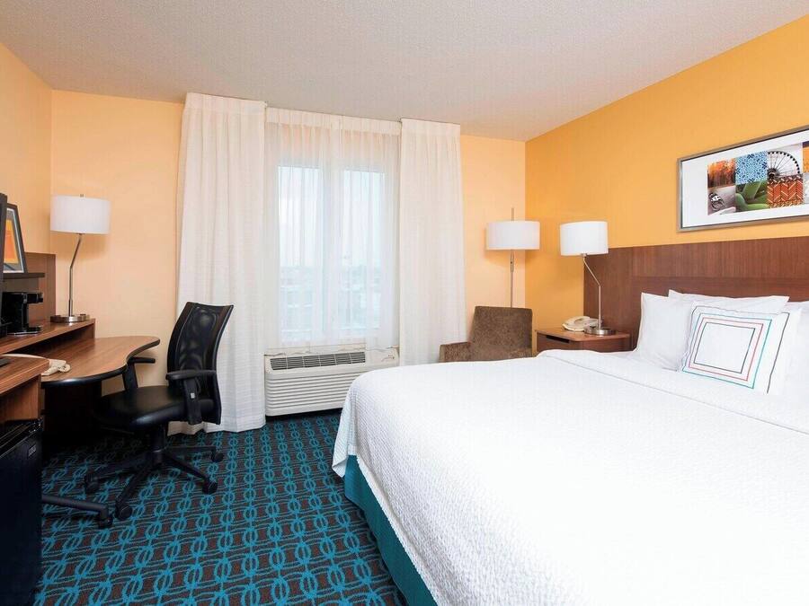 Quarto de hotel com cama, escrivaninha com cadeira, televisão e janela com cortinas.