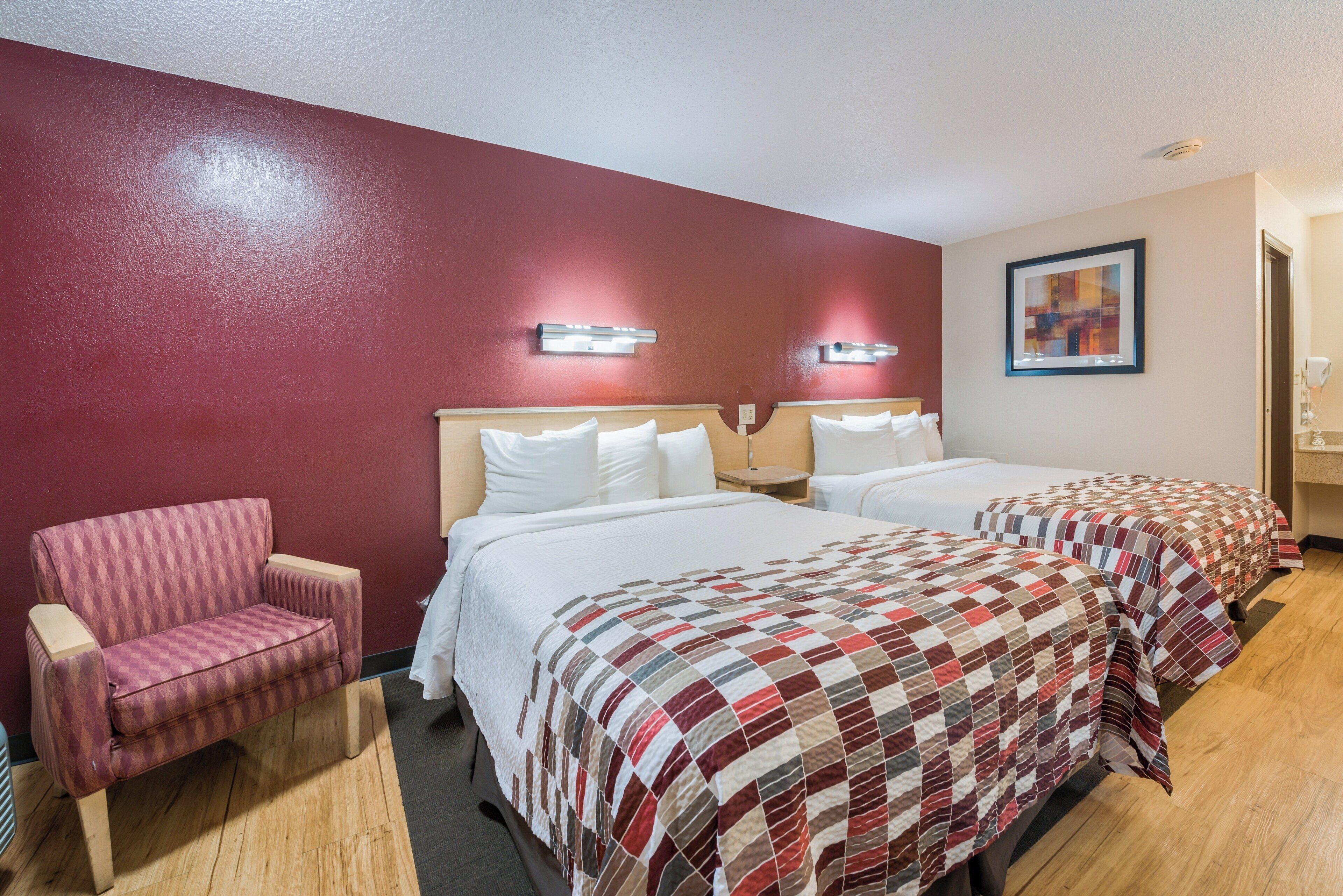 Quarto de hotel com duas camas, uma parede vermelha, uma poltrona e um quadro.