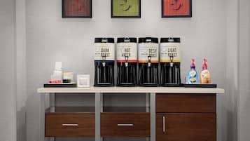 Uma estação de café com quatro dispensadores identificados como: torra escura, água quente, descafeinado e torra clara. Há também duas canecas e dois frascos de leite na bancada.