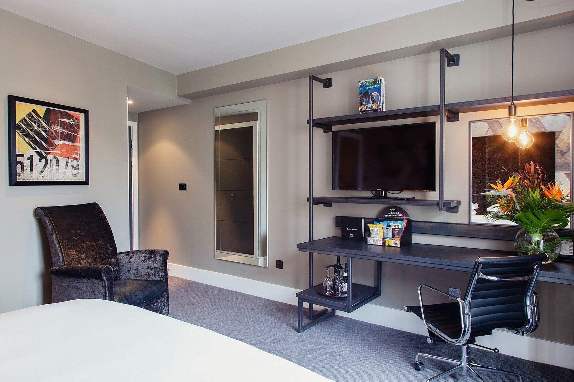 Quarto de hotel moderno com uma cama, uma mesa com televisão de tela plana, uma cadeira, uma estante de livros e um espelho grande.