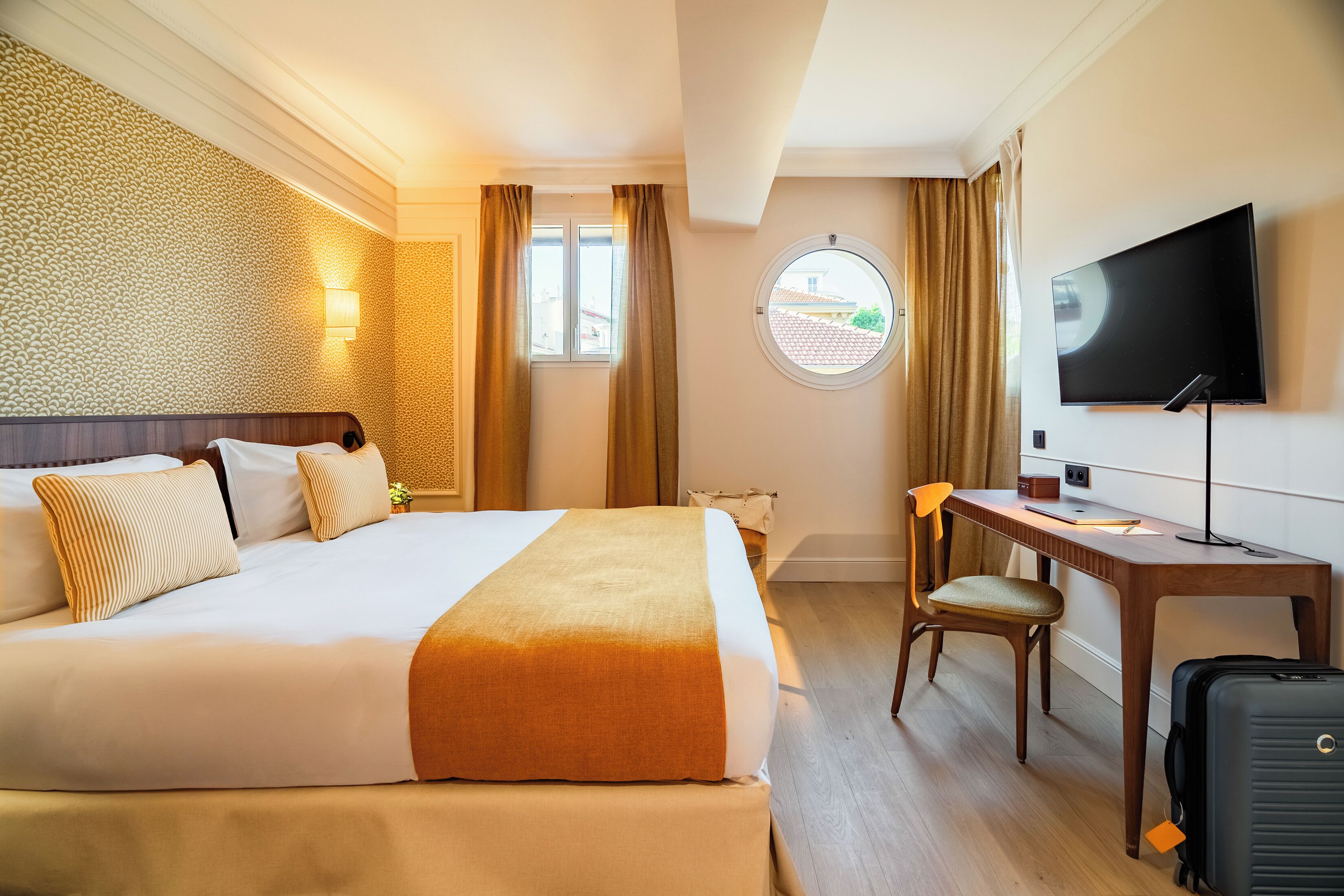 Quarto de hotel com uma cama grande, uma escrivaninha, uma cadeira, uma televisão e uma mala.