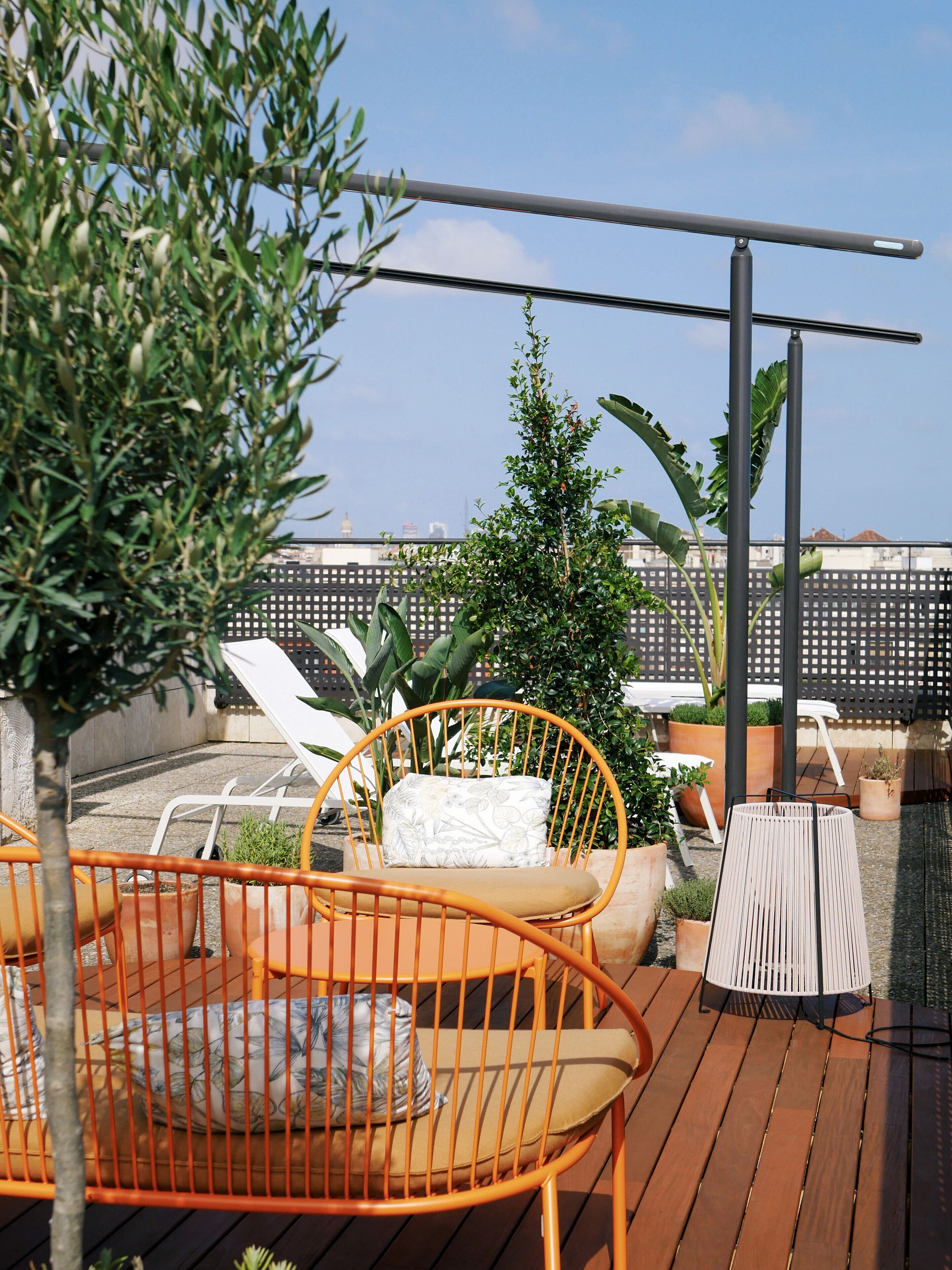 Terraço no telhado com deck de madeira, cadeiras de metal laranja, mesa branca e plantas em vasos.