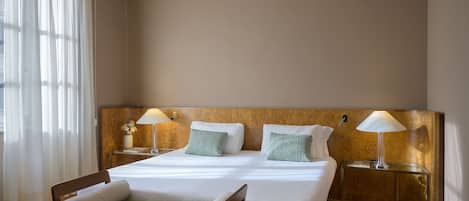 Quarto de hotel com uma cama grande, duas mesas de cabeceira com luminárias, uma cabeceira de madeira e uma janela com cortinas.