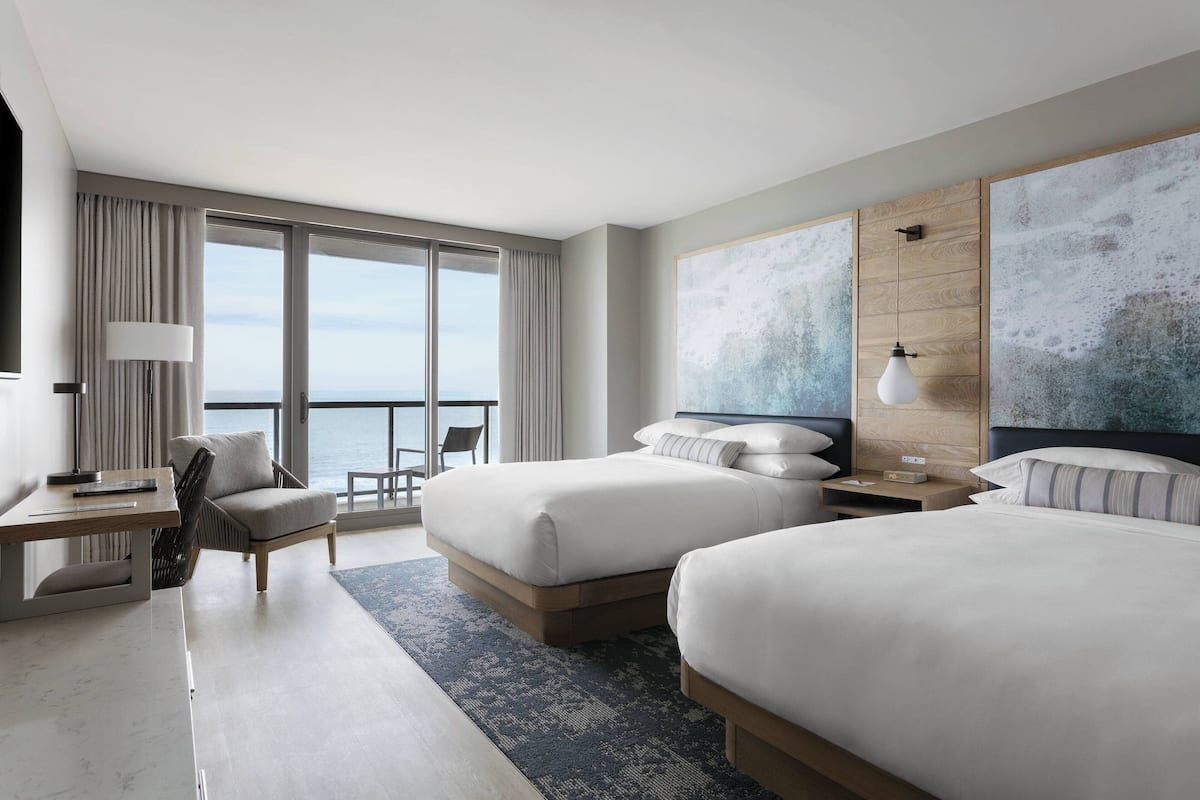 Quarto de hotel com duas camas, uma escrivaninha, uma cadeira e uma janela grande com vista para o mar.