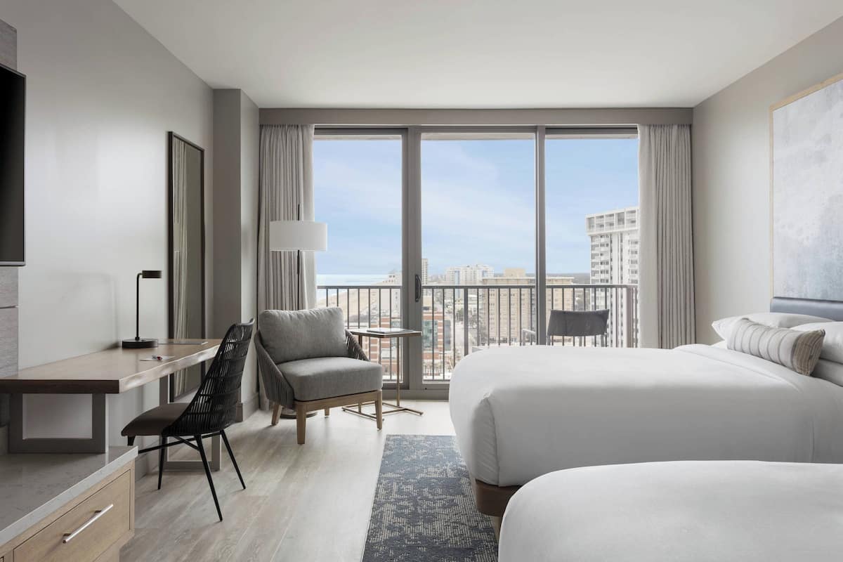 Quarto de hotel com uma cama grande, uma escrivaninha, uma cadeira e vista da cidade por amplas janelas.