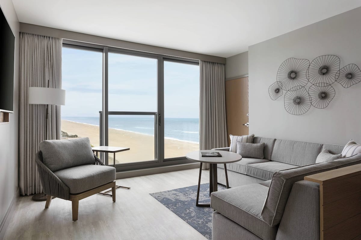 Quarto de hotel moderno com uma grande janela que oferece vista para a praia, um sofá cinza, uma mesa pequena e um detalhe decorativo na parede.