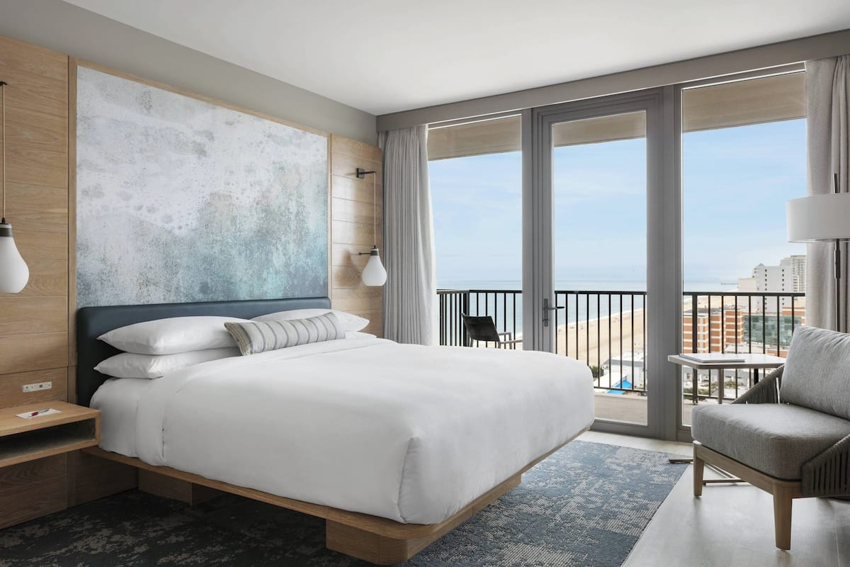 Quarto de hotel moderno com uma cama grande, vista para a praia e varanda com uma cadeira.