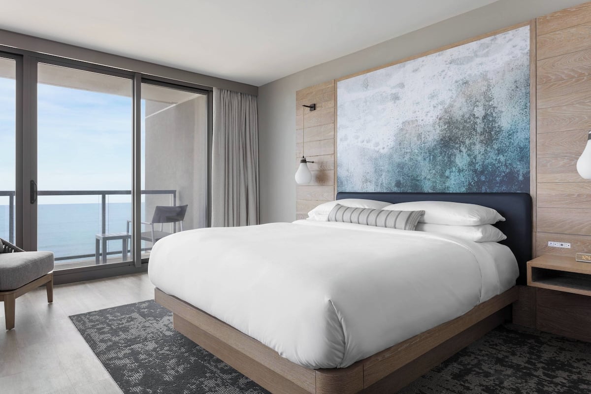 Quarto de hotel moderno com uma cama grande, vista para o mar, varanda e cabeceira de madeira.