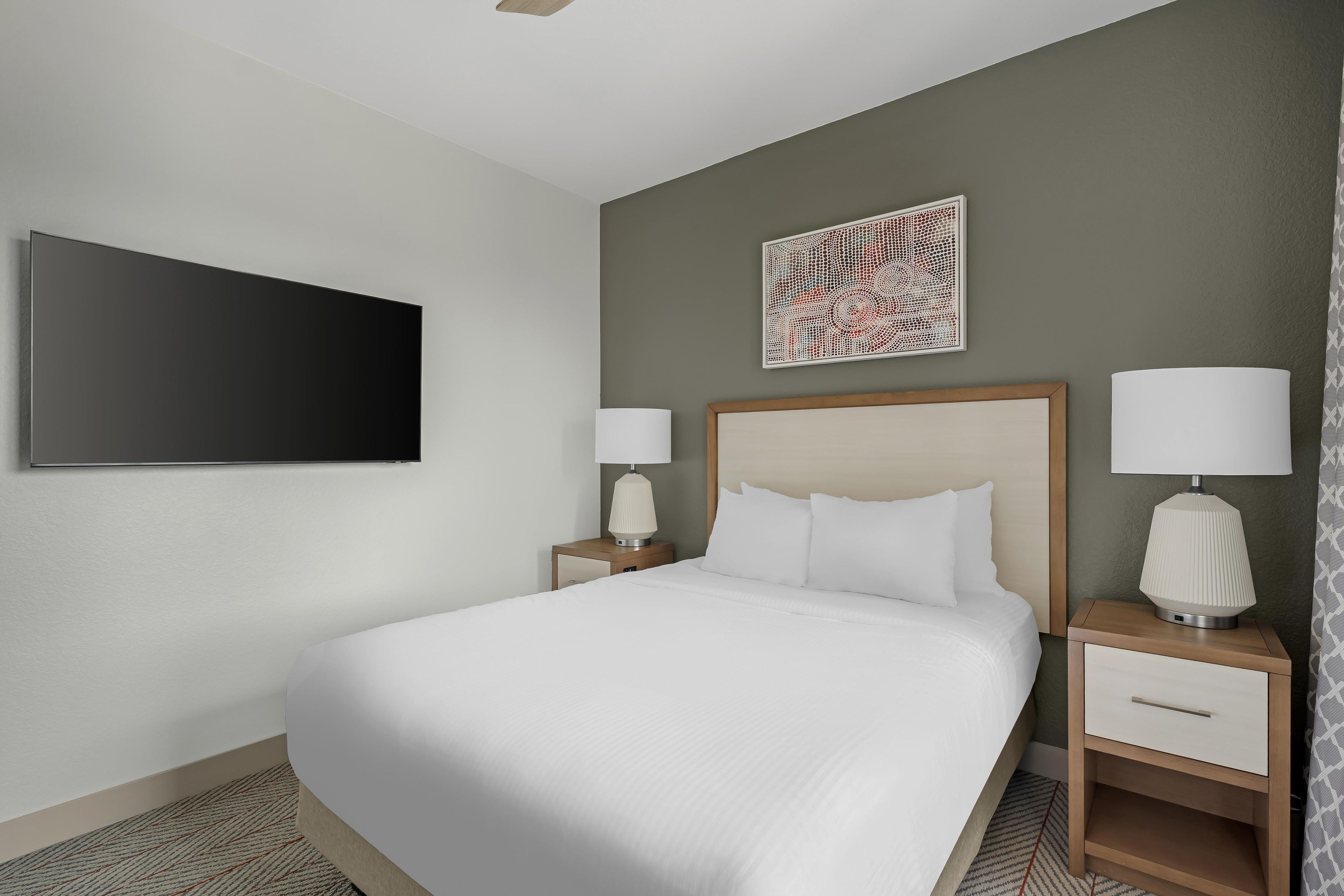 Quarto de hotel com uma cama grande, duas mesas de cabeceira com abajures, uma TV de tela plana e um painel decorativo na parede.