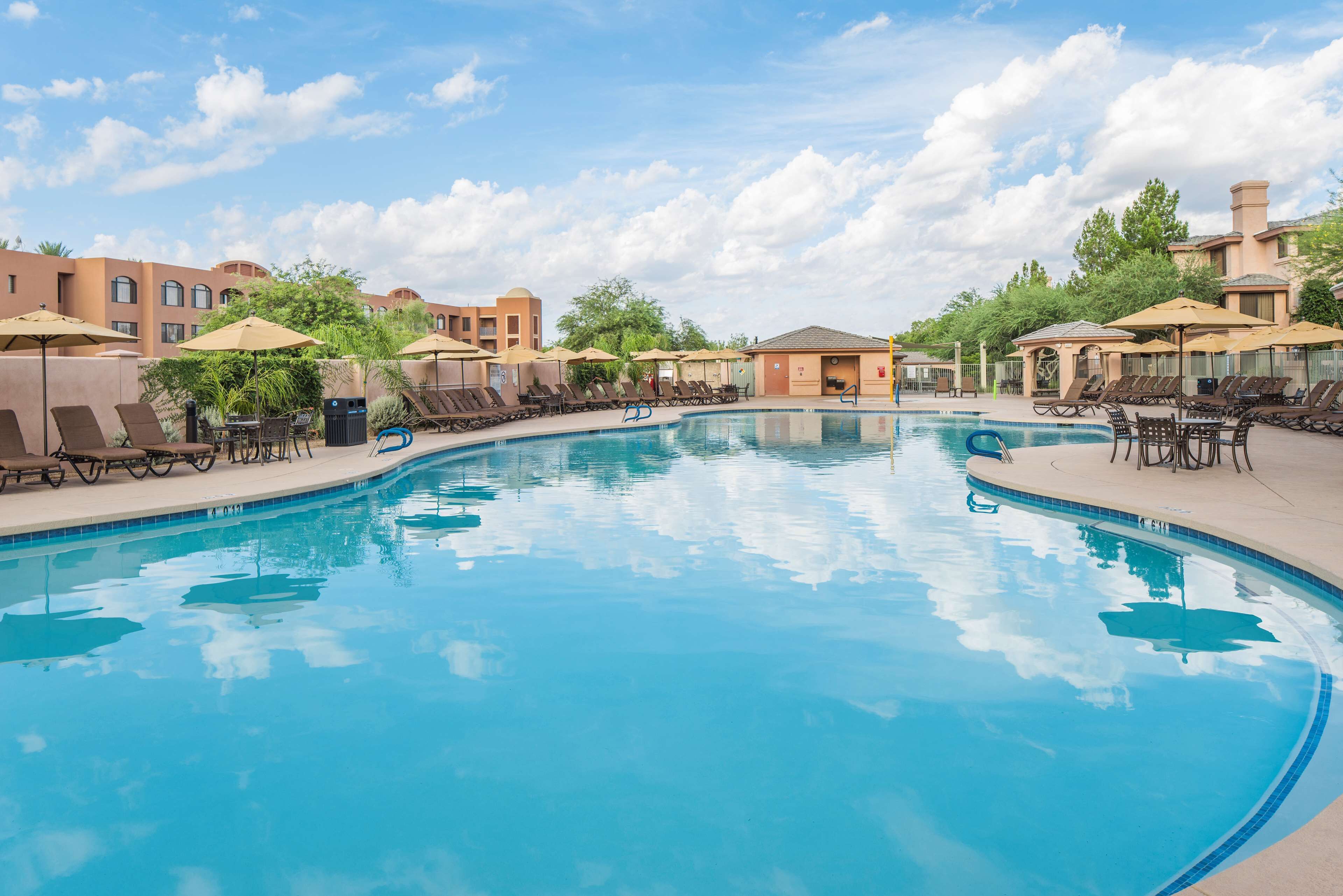 Piscina com espreguiçadeiras e guarda-sóis, rodeada por edifícios sob um céu claro.