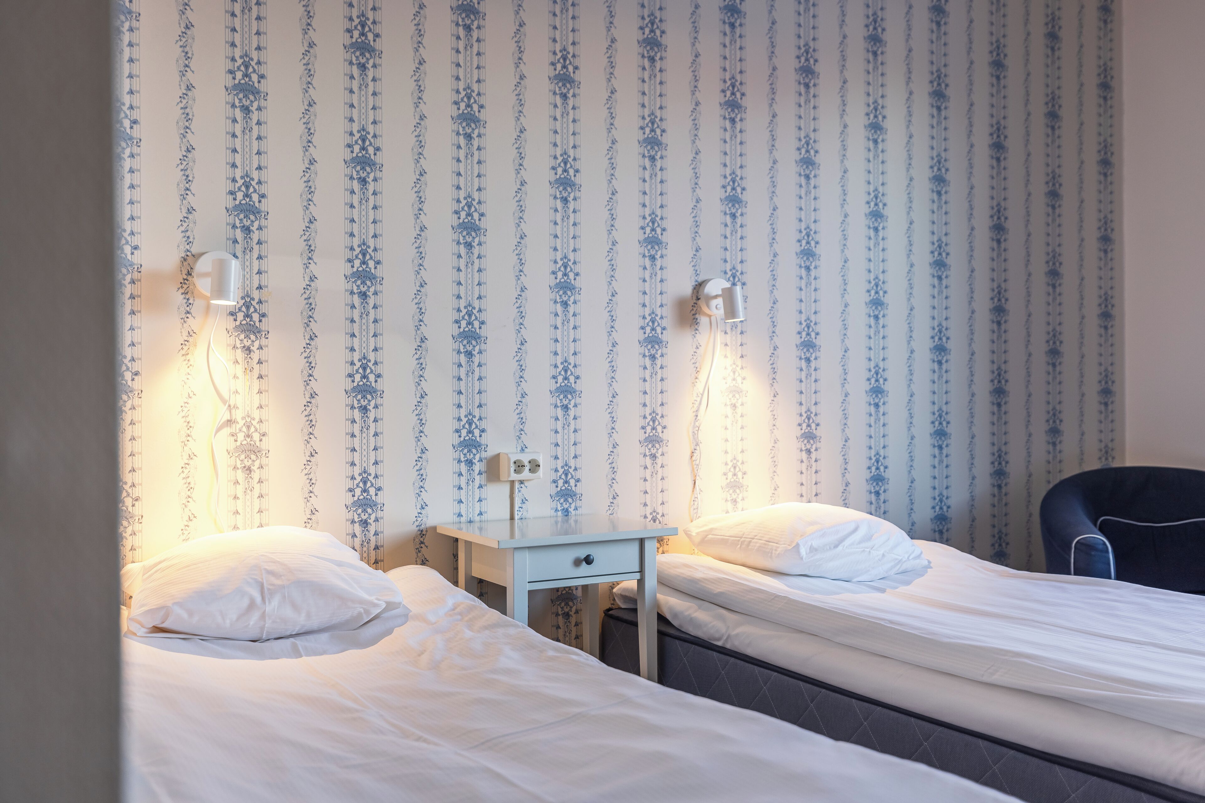 Quarto de hotel com duas camas de solteiro, uma mesinha de cabeceira com abajur e uma cadeira.