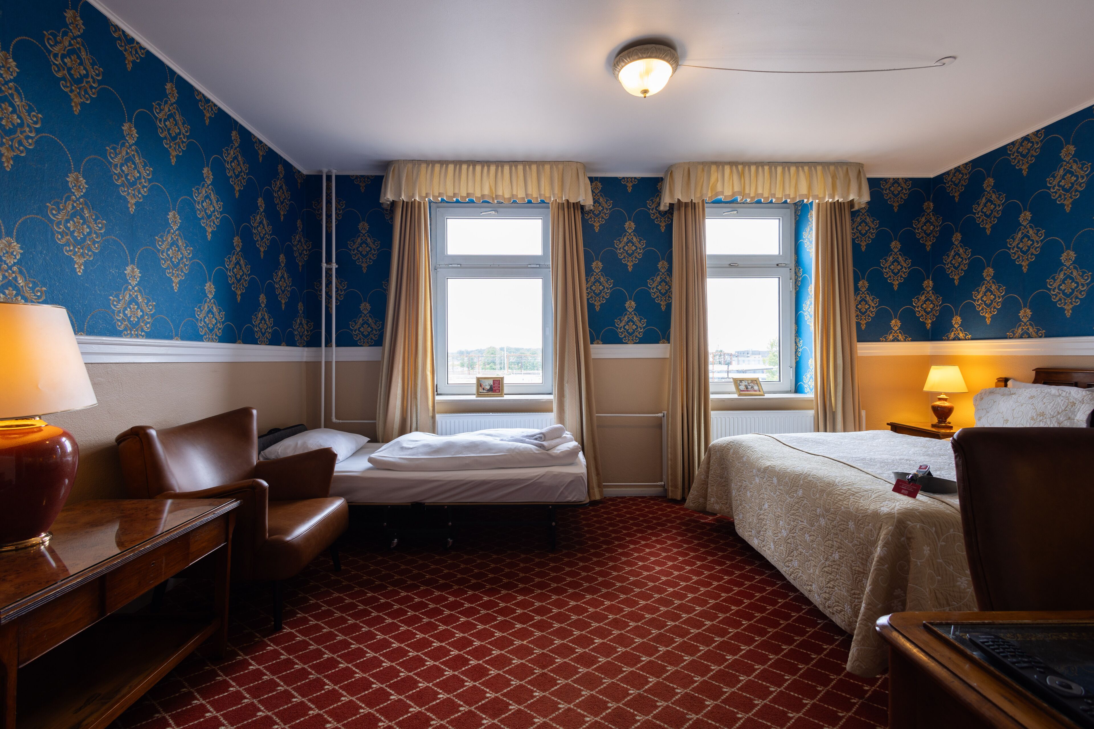 Quarto de hotel com duas camas, uma escrivaninha, uma cadeira e uma janela com cortinas.