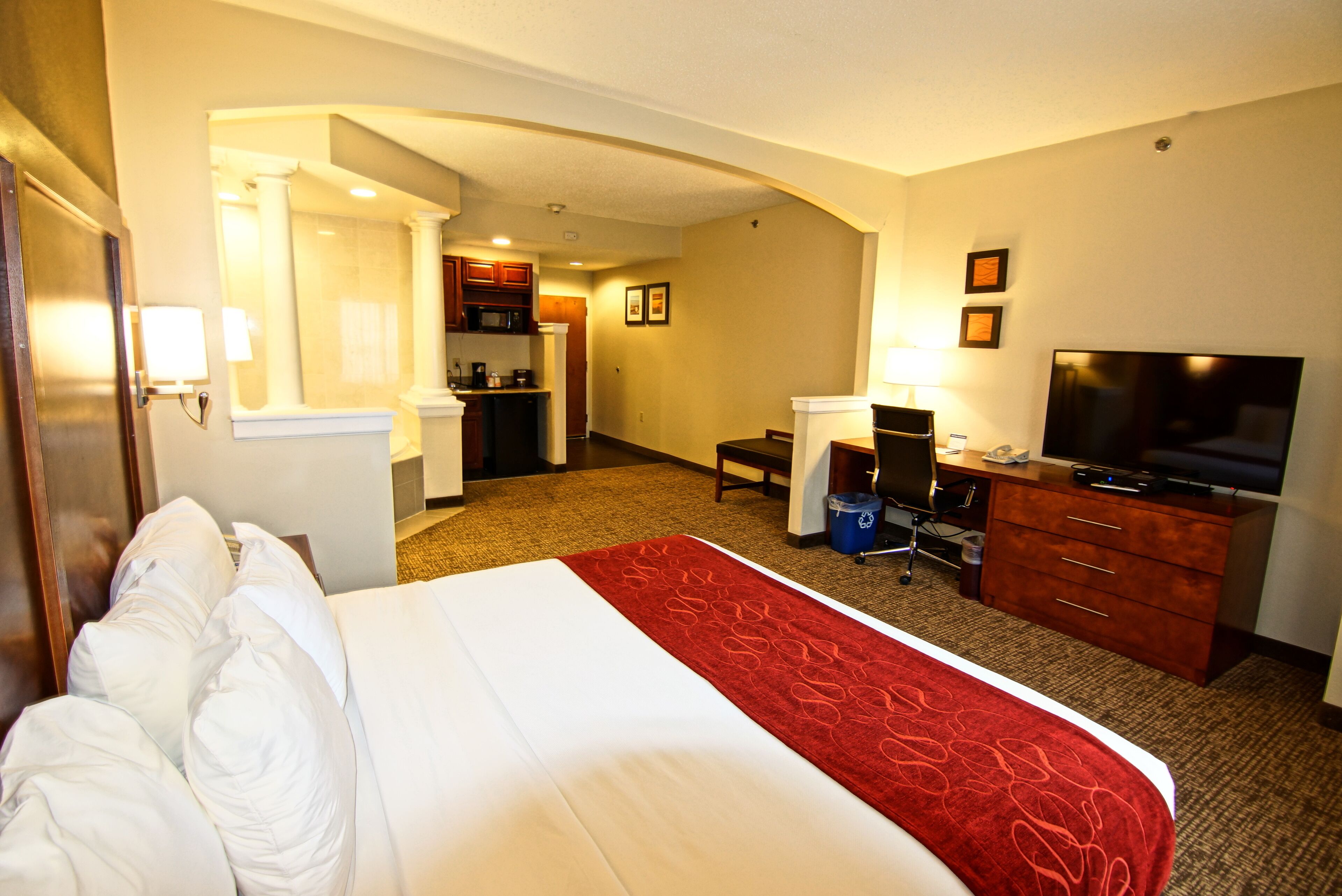 Quarto de hotel com uma cama grande, uma escrivaninha com computador, televisão e um espaço com cozinha.
