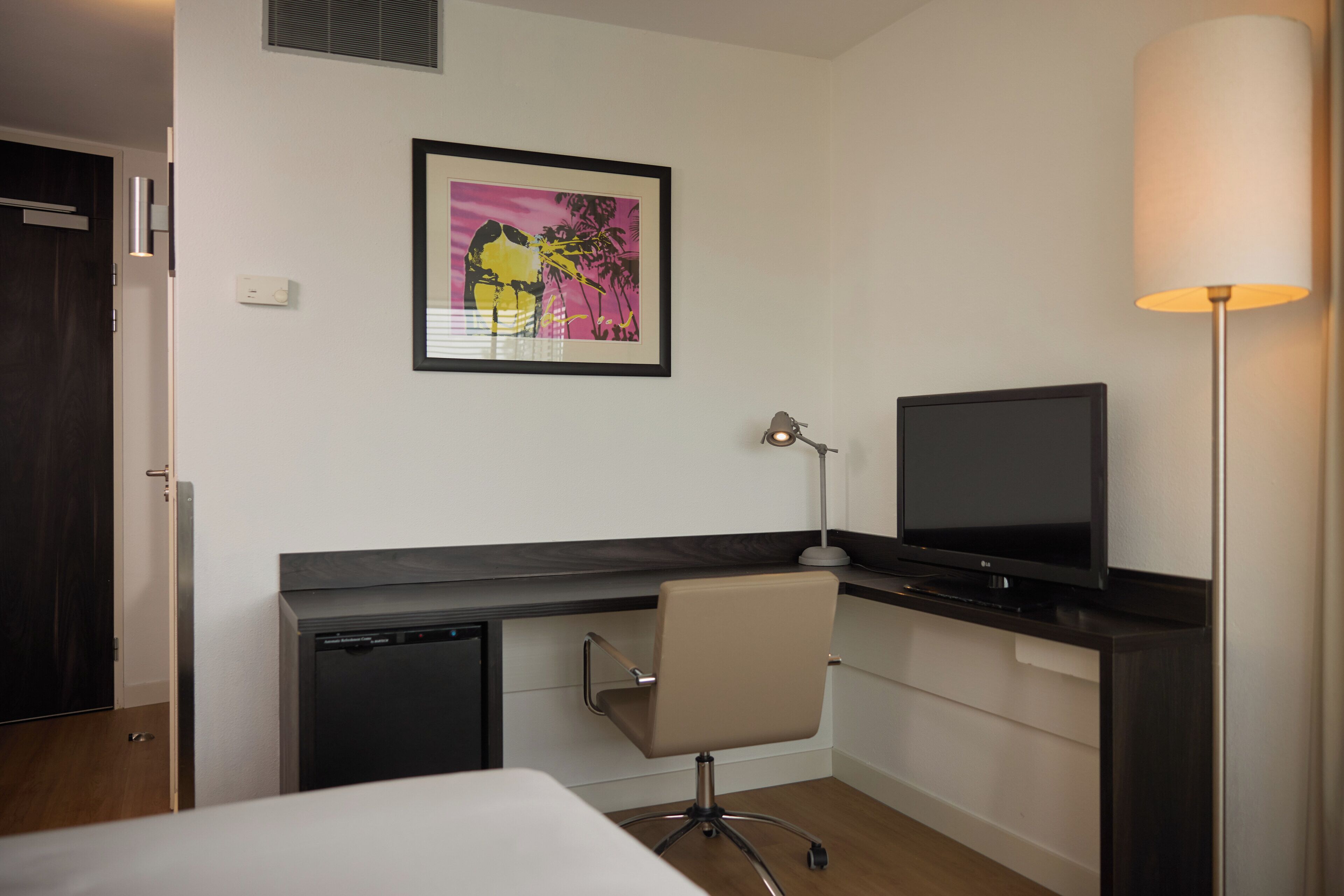 Quarto de hotel com cama, secretária com monitor de computador, cadeira, luminária de secretária e televisão presa à parede.