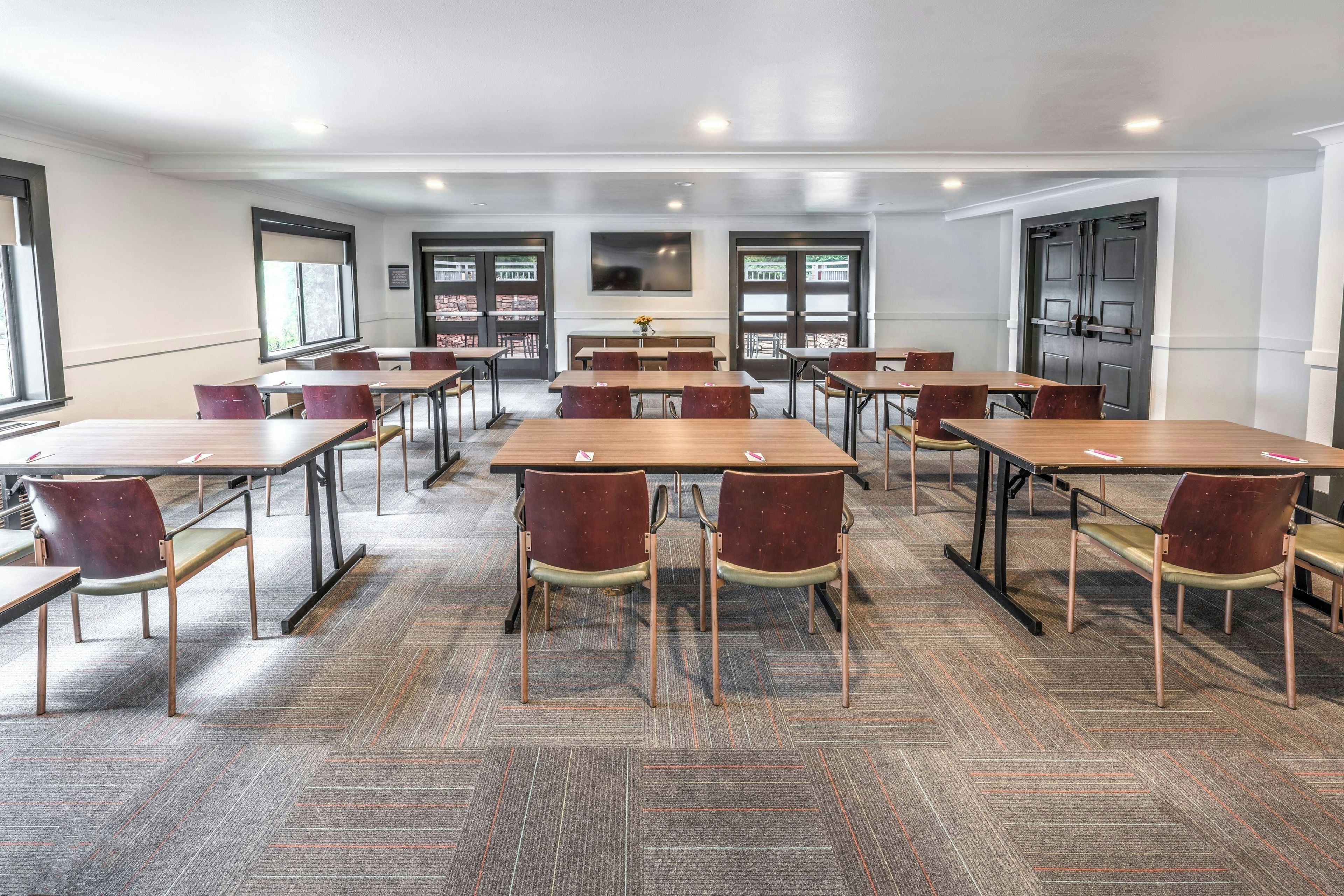 Sala de conferência com várias mesas e cadeiras, piso coberto de carpete e grandes janelas.