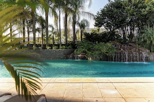 Piscina com cachoeira, rodeada por vegetação tropical e um muro de pedra.