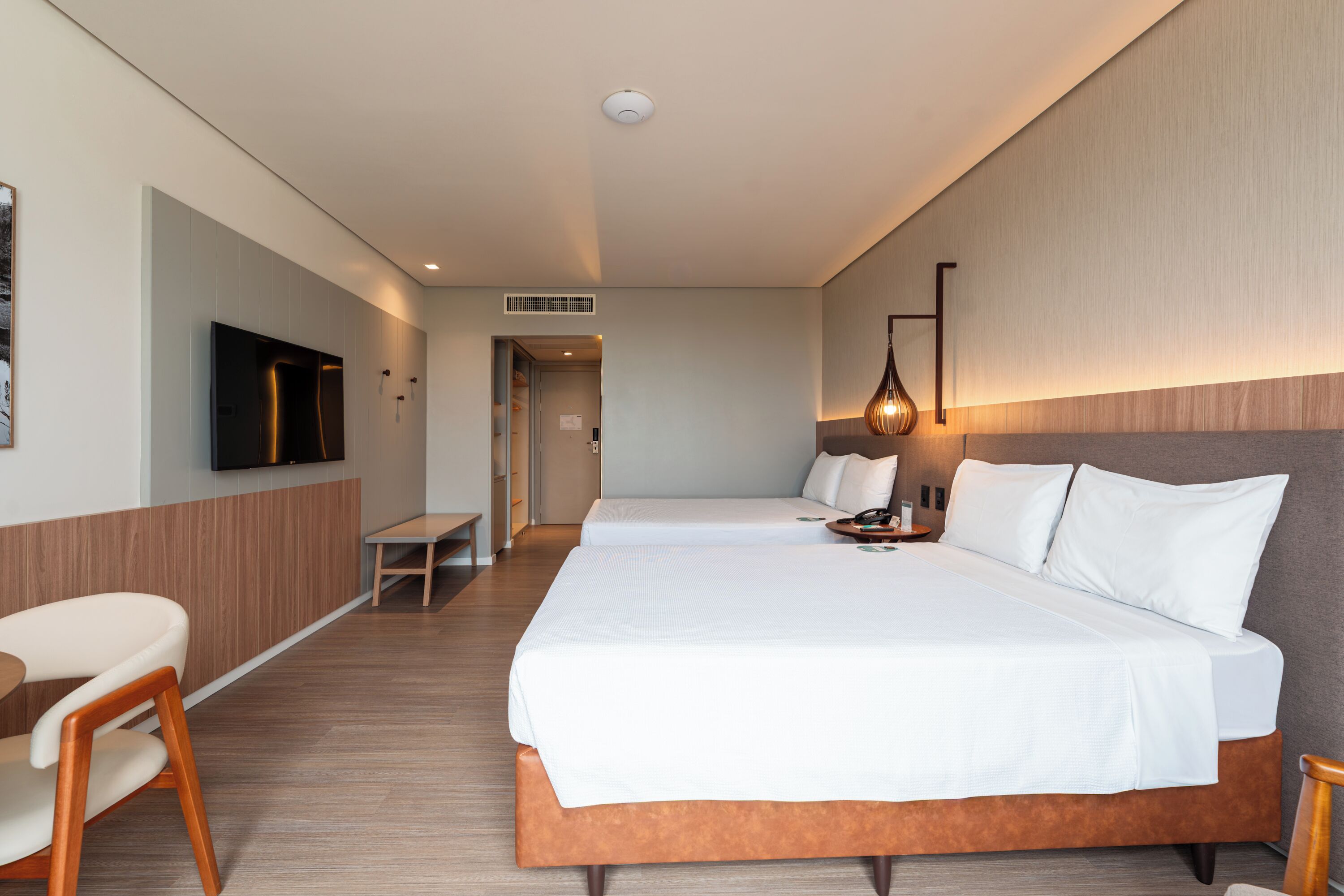 Quarto de hotel com duas camas, uma mesa, uma cadeira, uma televisão e um abajur.