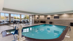 Piscina interior com paredes de vidro transparente, área de descanso com cadeiras e mesas, e vista para carros estacionados do lado de fora.