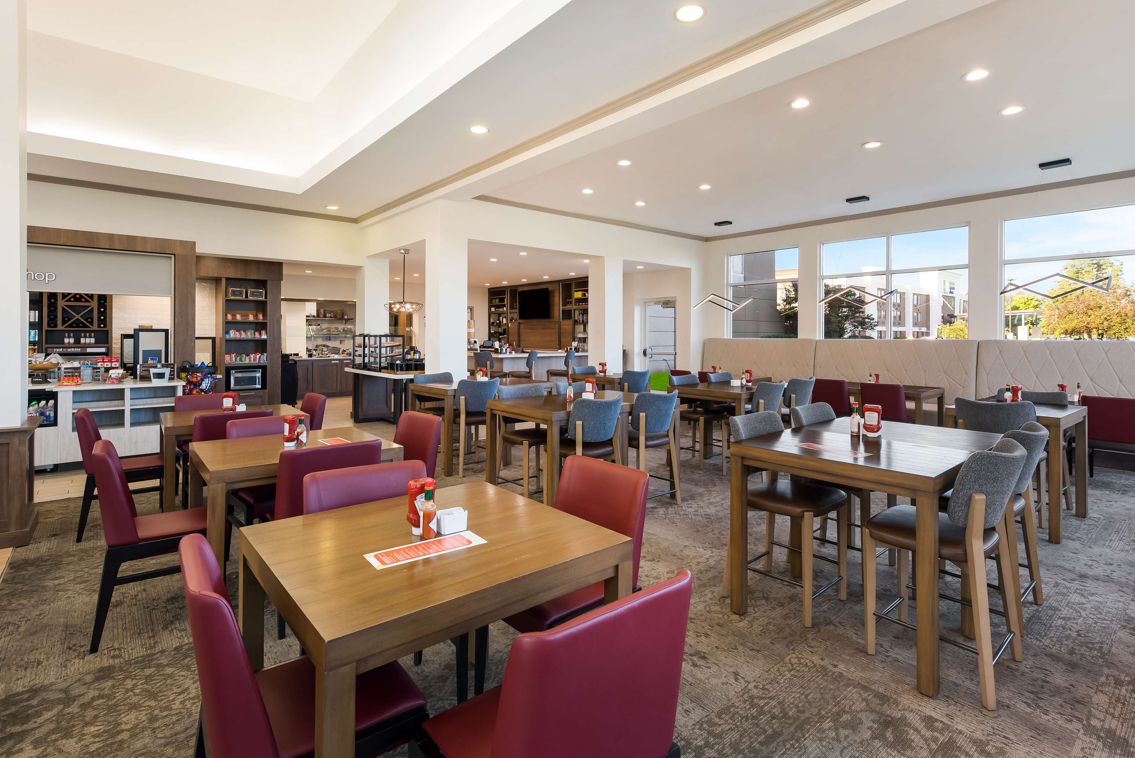Um restaurante moderno com várias mesas e cadeiras, área de cozinha visível e grandes janelas com vista para o exterior.