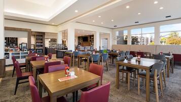 Um restaurante moderno com várias mesas e cadeiras, área de cozinha visível e grandes janelas com vista para o exterior.