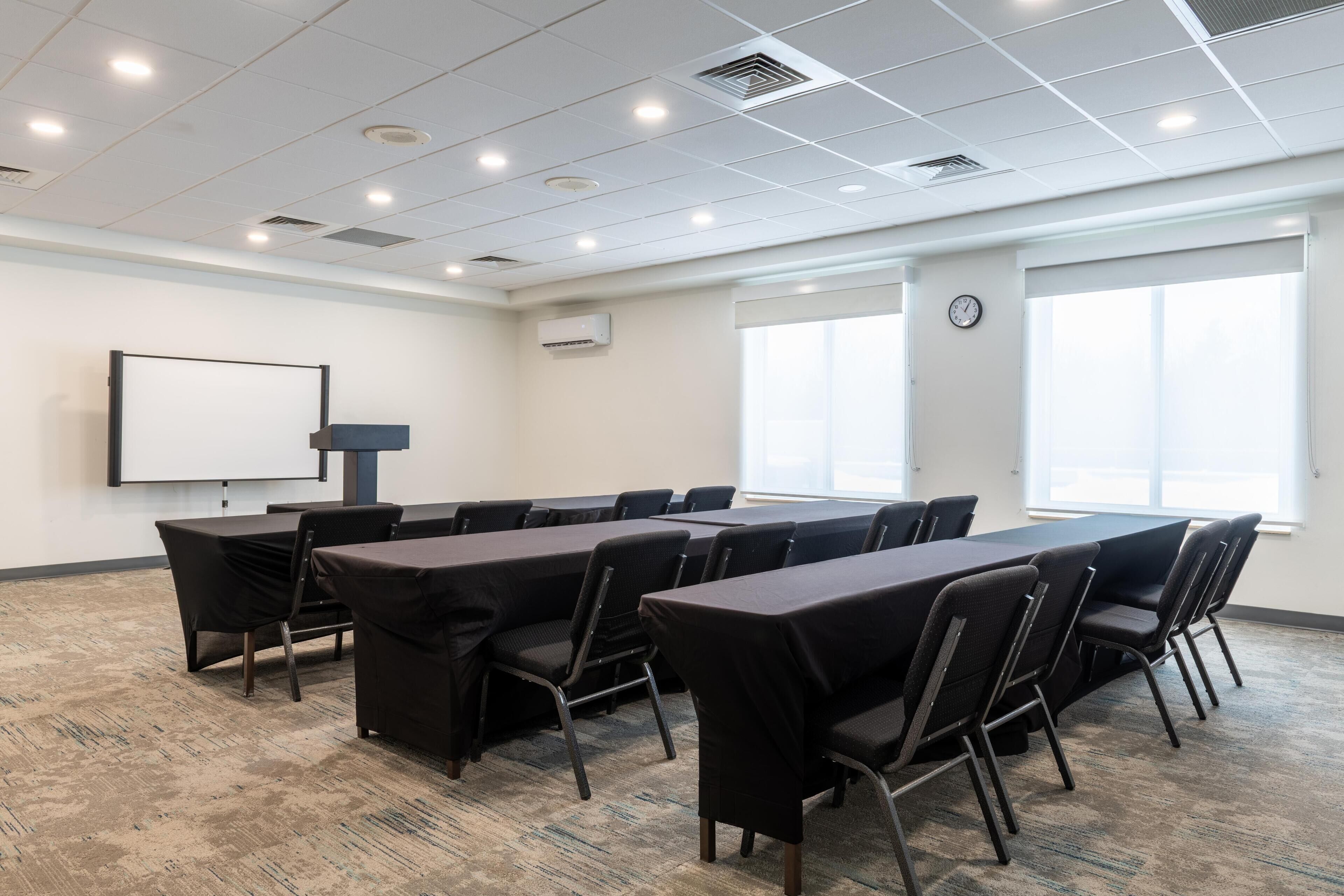 Uma sala de conferências com uma mesa grande, cadeiras, uma tela de projetor e um relógio na parede.