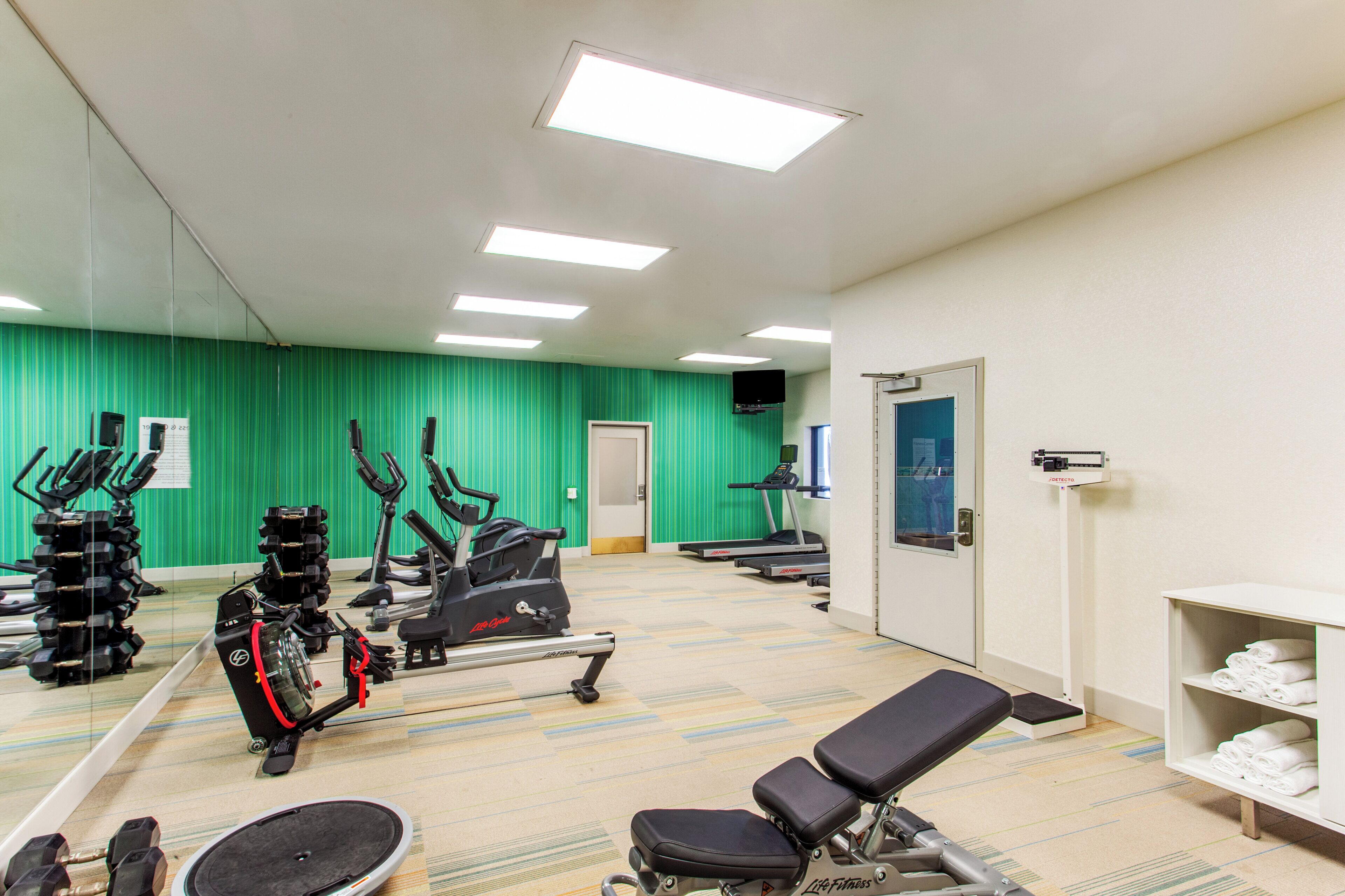 Uma academia com equipamentos de ginástica, um espelho e uma porta que dá para outro cômodo.