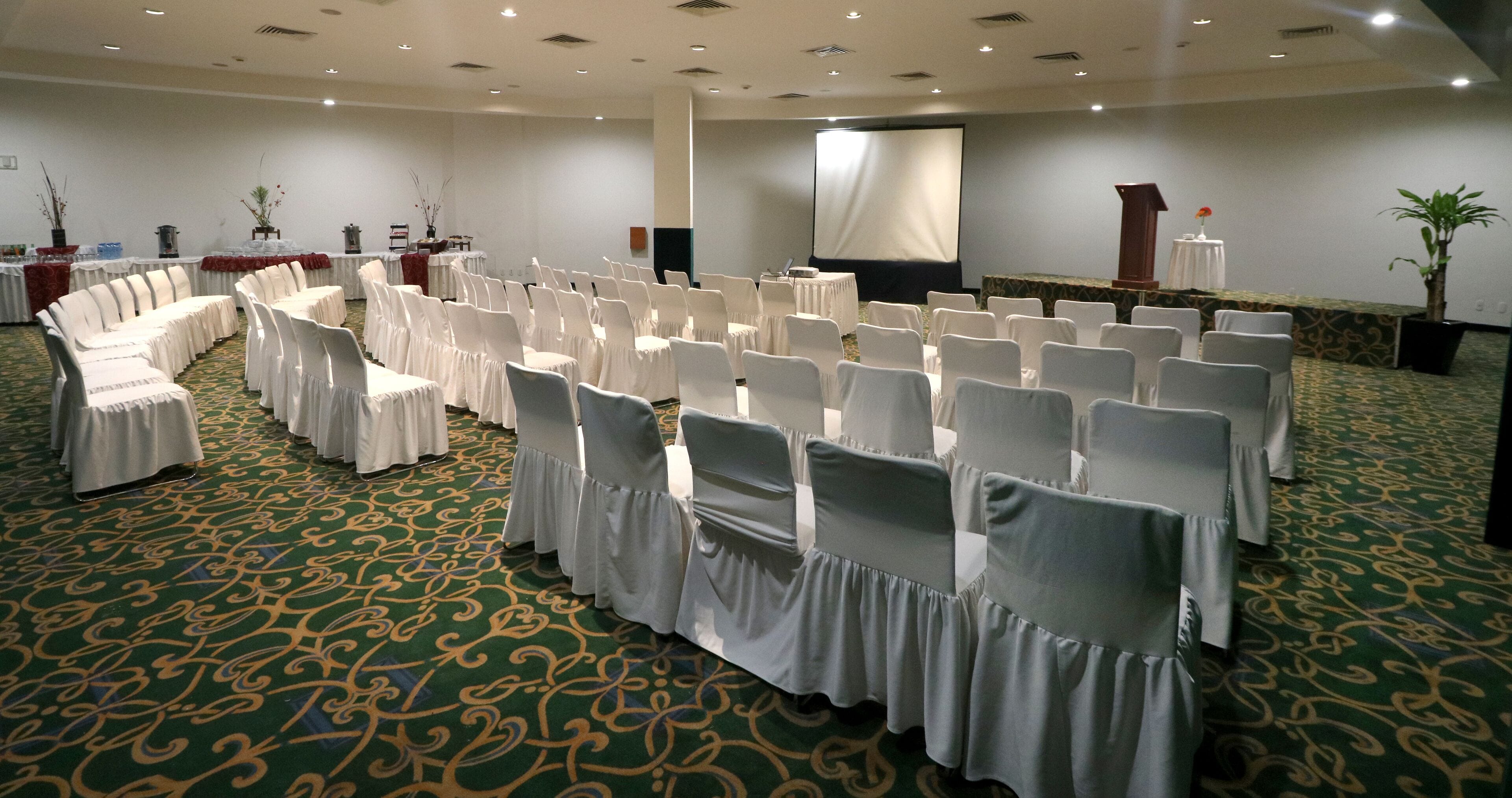Sala de conferência com cadeiras dispostas em fileiras, um púlpito e uma tela.