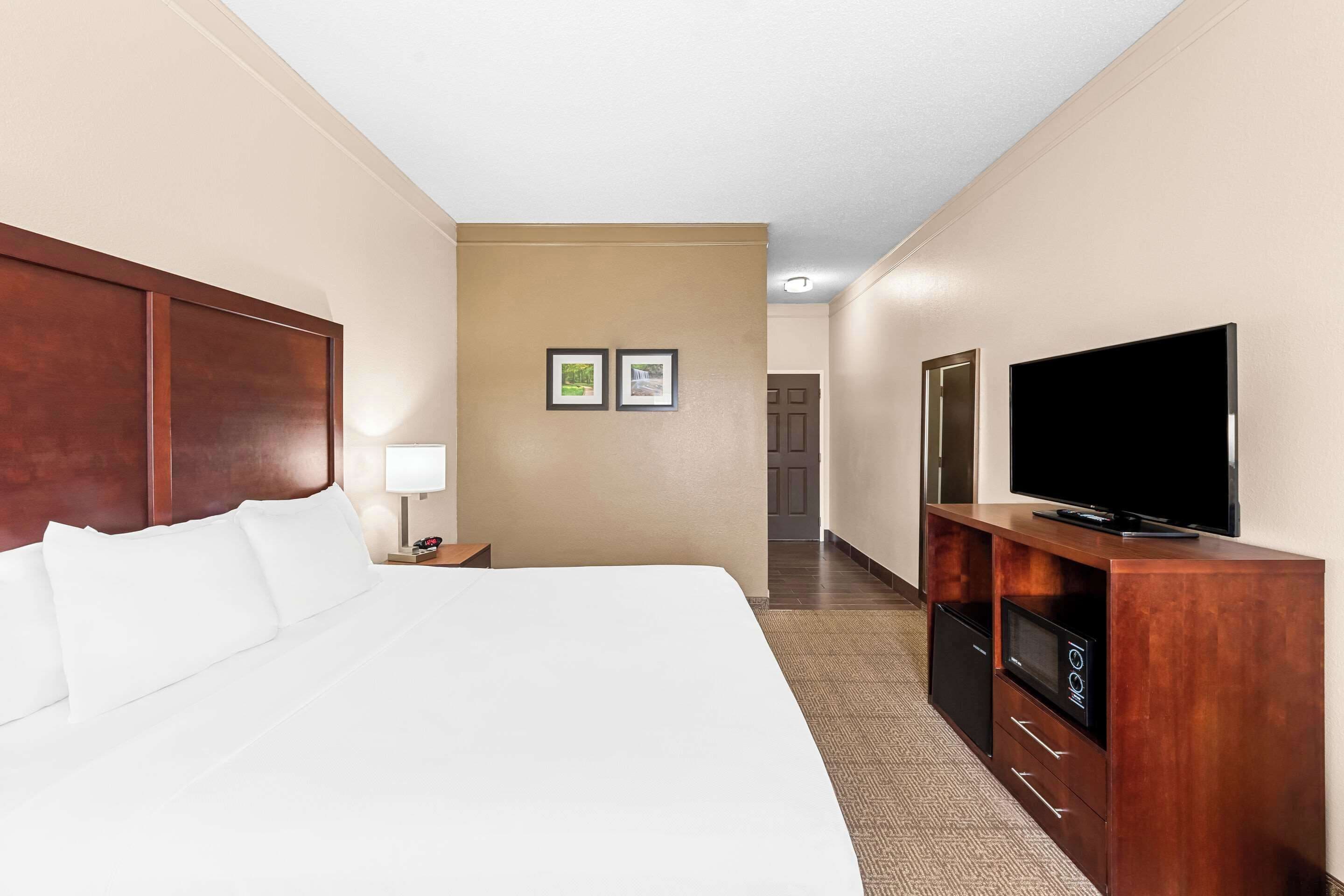 Quarto de hotel com uma cama grande, uma TV sobre um suporte de madeira e uma pequena escrivaninha com uma luminária.