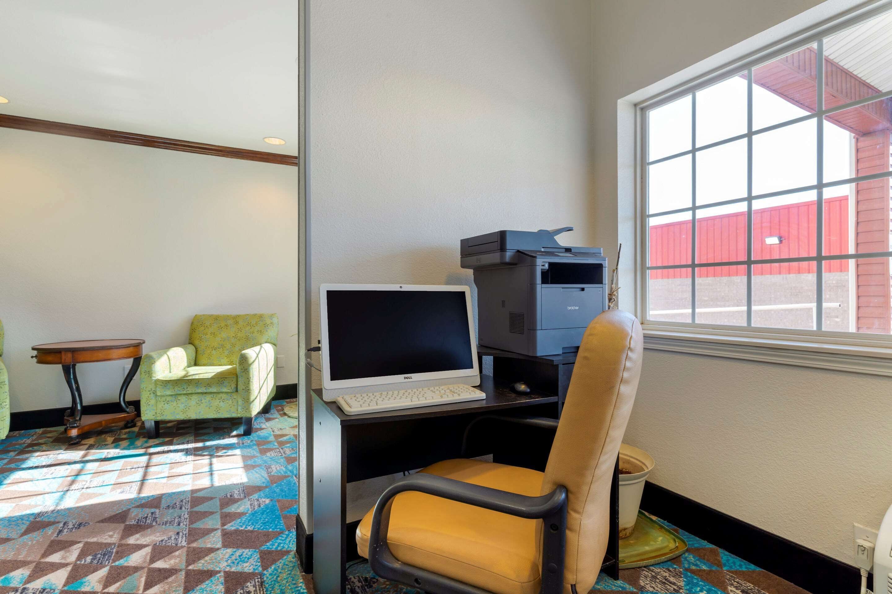 Quarto de hotel com uma escrivaninha, um computador, uma impressora, uma janela com vista para um edifício vermelho, um tapete estampado e uma poltrona verde.