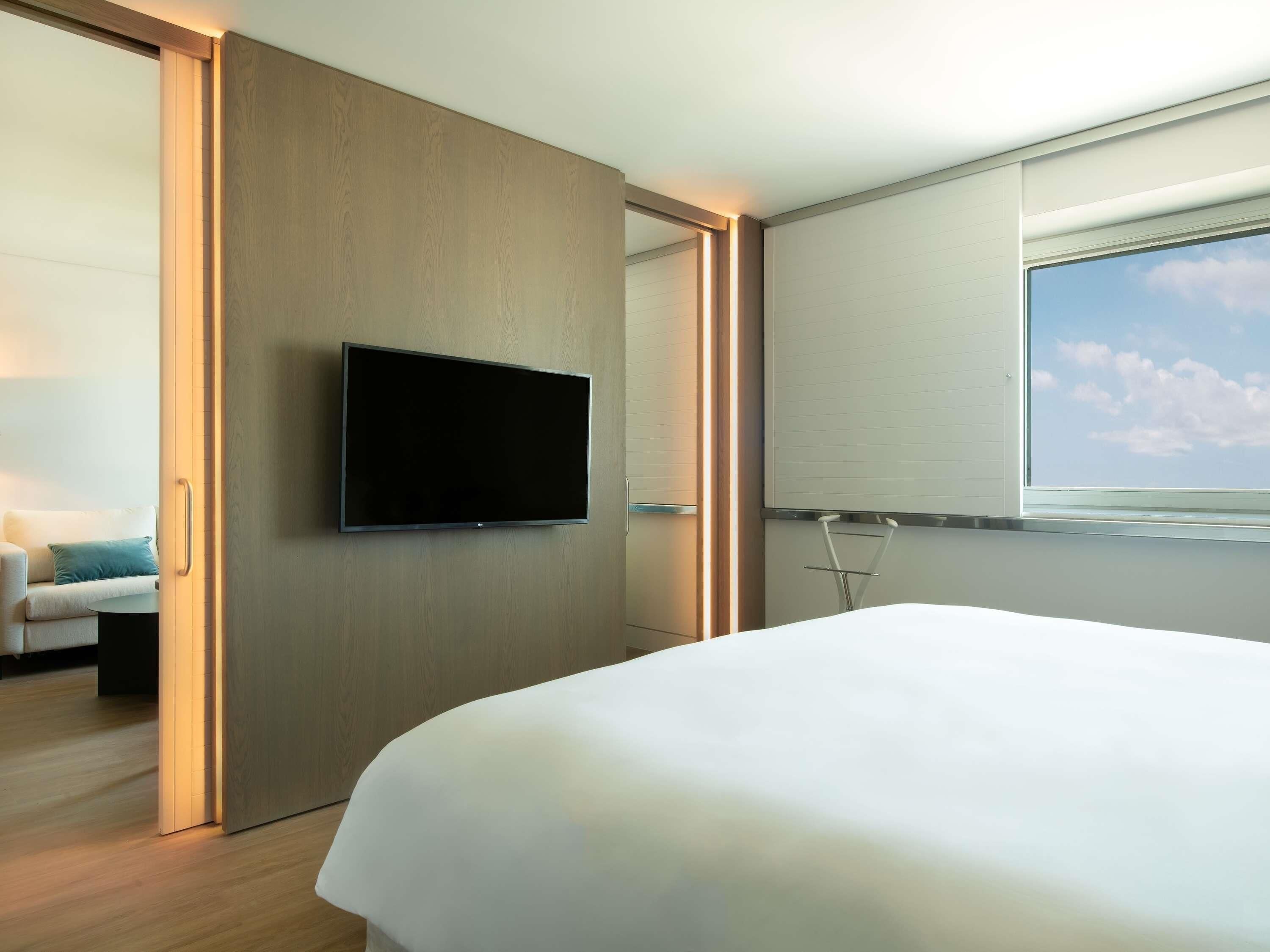 Quarto de hotel moderno com uma cama grande, televisão de tela plana fixada em um painel de madeira, janela com vista para o céu e um pequeno espaço de estar com sofá e mesa.