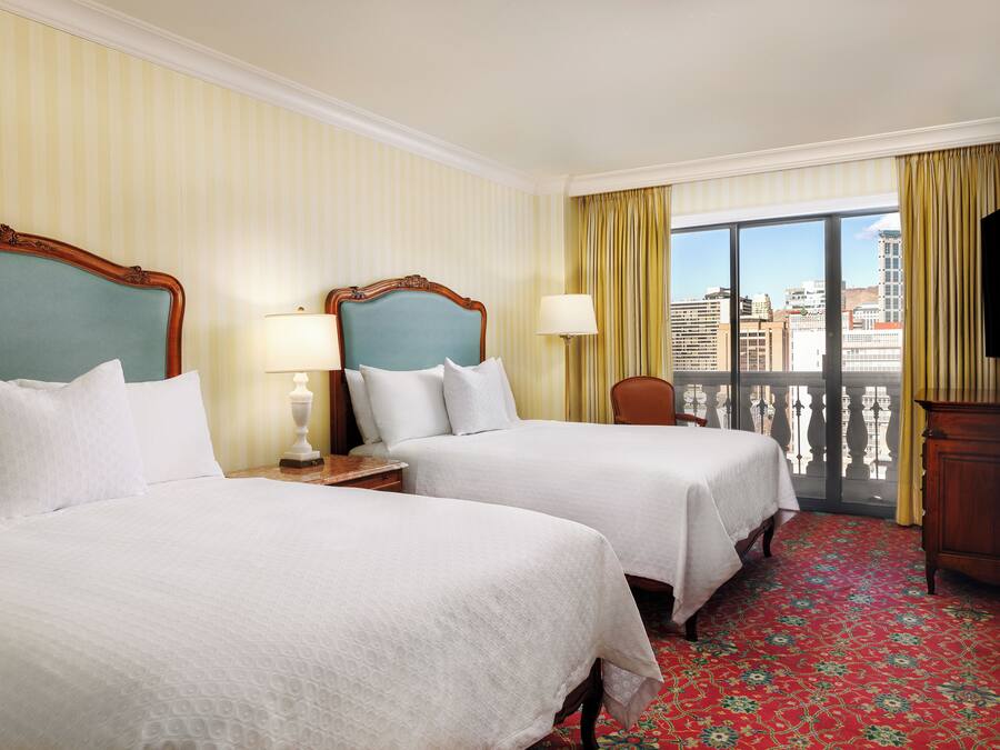 Quarto de hotel com duas camas, uma janela grande com vista para a cidade, televisão e um armário de madeira.