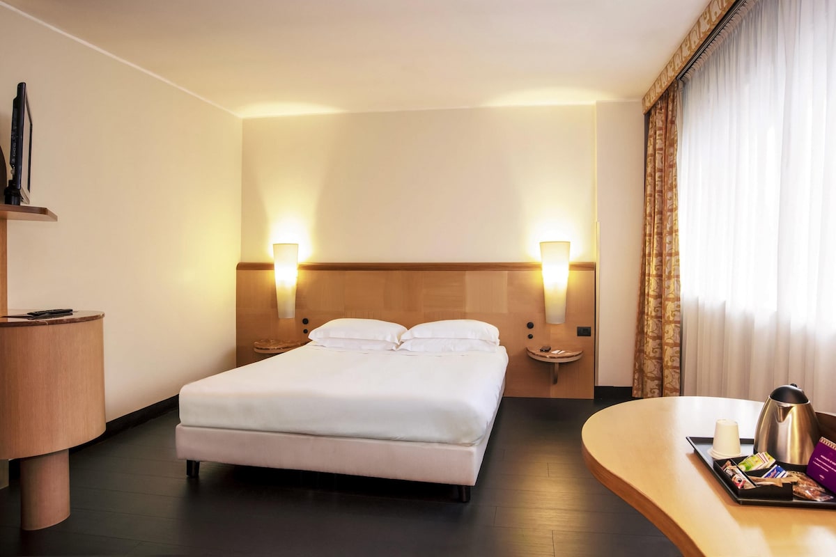 Quarto de hotel com cama, mesas de cabeceira, escrivaninha com bandeja de objetos e janela com cortinas.