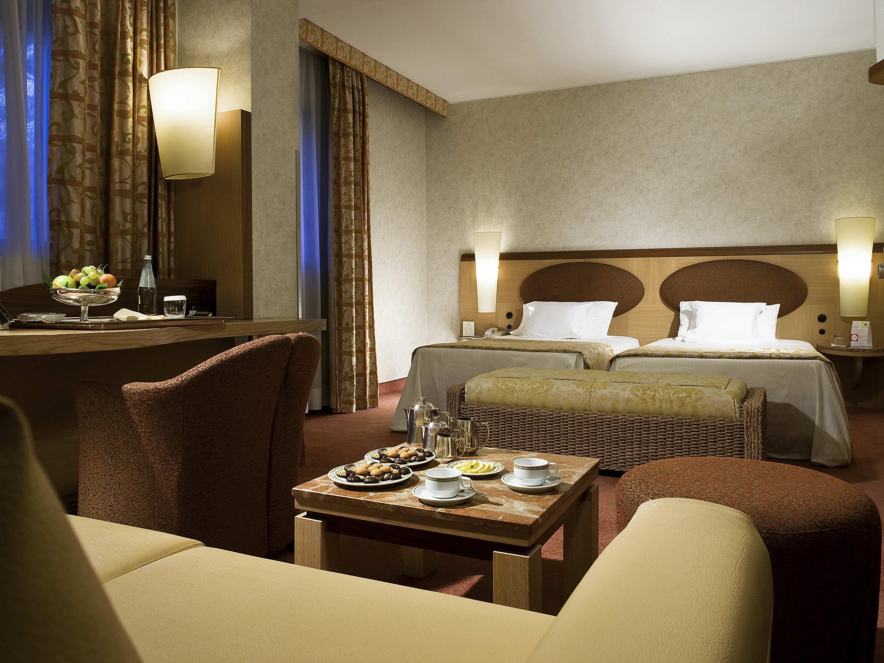 Quarto de hotel com cama, duas mesas de cabeceira, escrivaninha com cadeira, uma pequena mesa com doces e uma cadeira de vime.