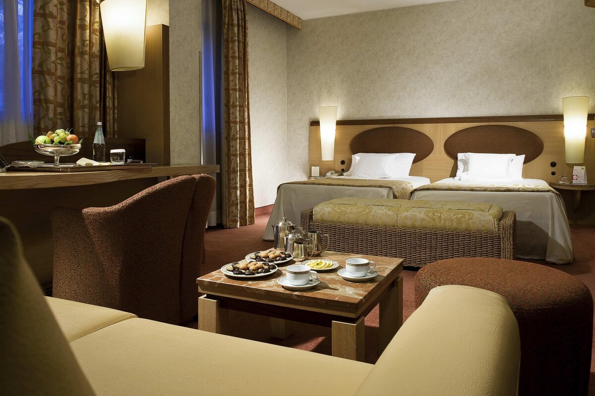 Quarto de hotel com cama, duas mesas de cabeceira, escrivaninha com cadeira, uma pequena mesa com doces e uma cadeira de vime.