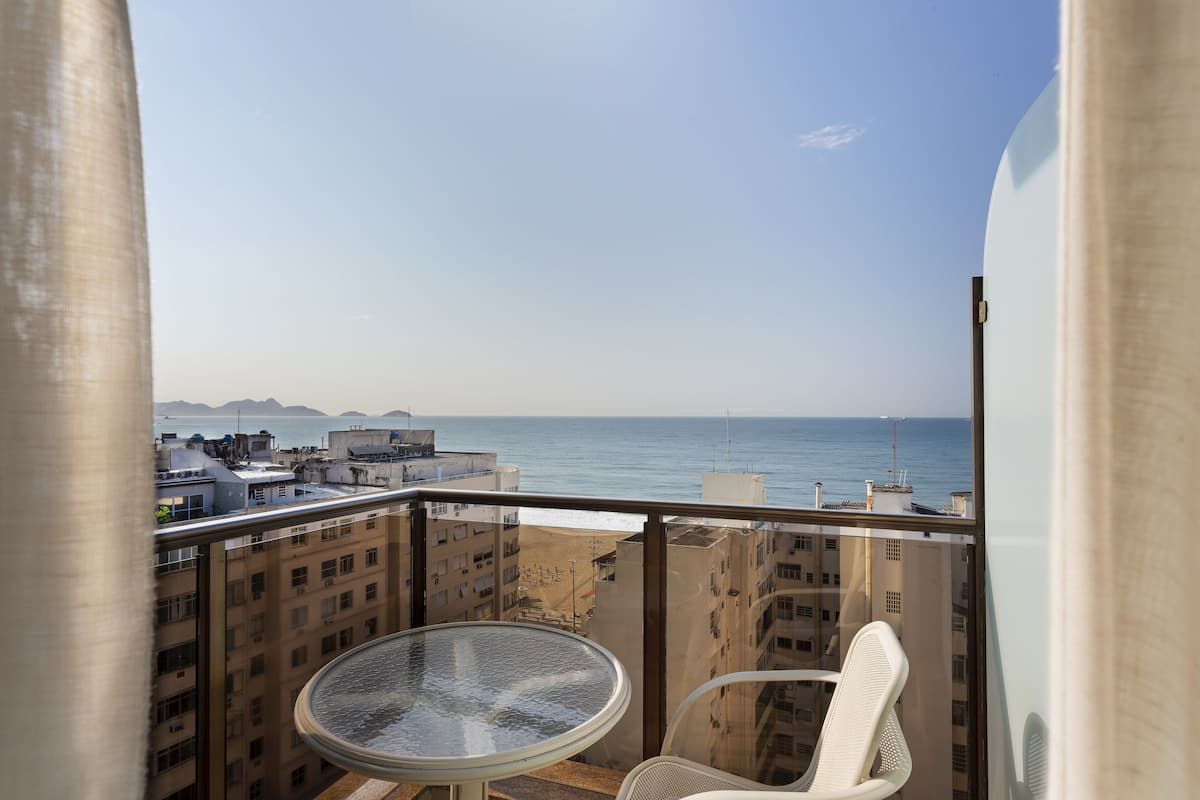 Varanda com mesa de vidro e uma cadeira, vista para a cidade e o mar.