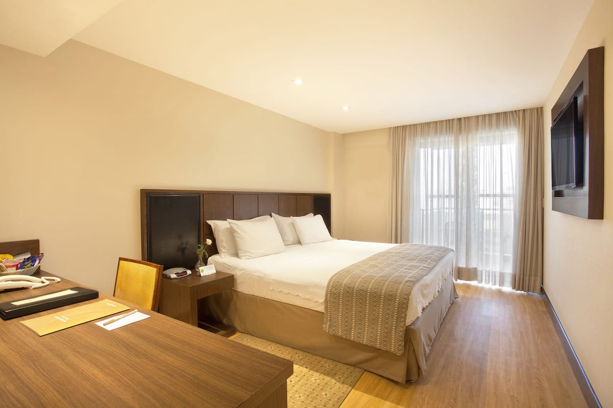 Quarto de hotel com uma cama grande, uma escrivaninha com telefone e televisão fixada na parede.