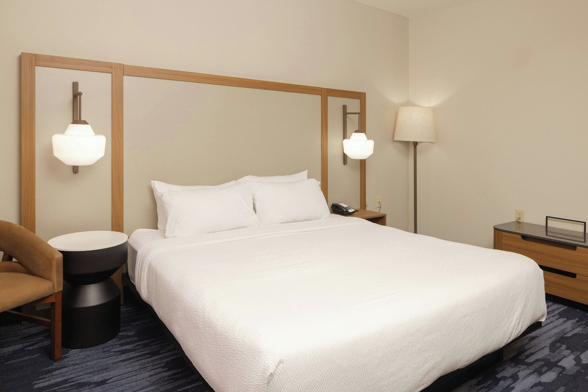 Quarto de hotel com uma cama grande, uma cadeira, uma mesinha de cabeceira, uma luminária e um ponto de luz na parede.