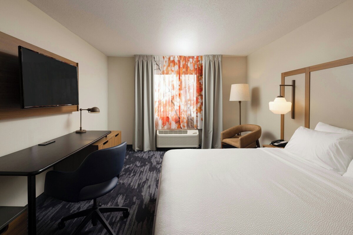 Quarto de hotel com cama, escrivaninha, cadeira, televisão, ar condicionado e luminária.