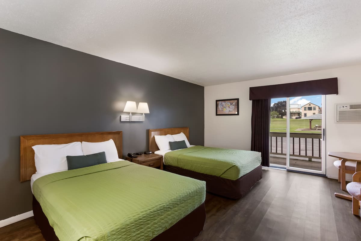 Quarto de hotel com duas camas, cabeceira de madeira, mesa de cabeceira com luminária, janela com vista para o exterior e porta de vidro deslizante.