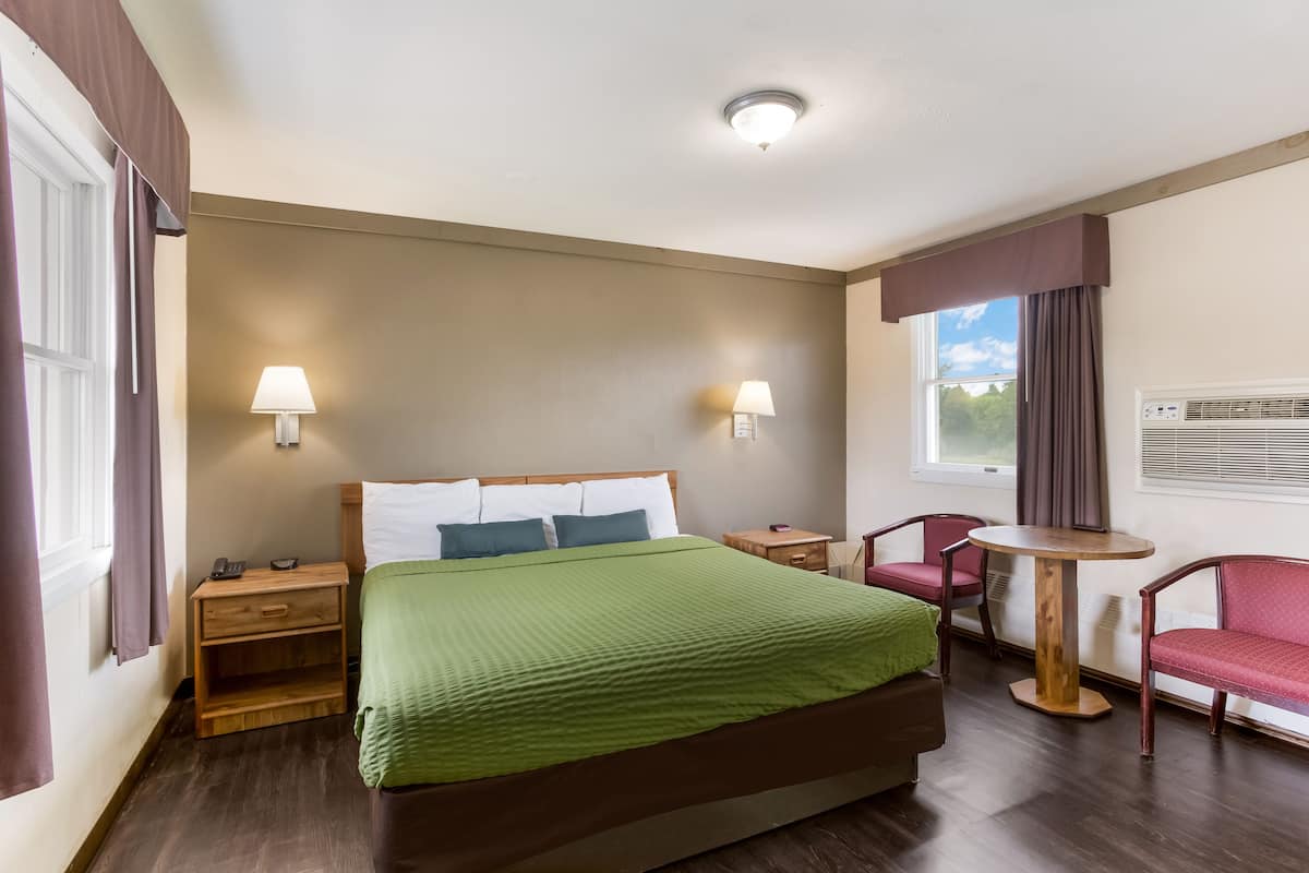 Quarto de hotel com cama, duas mesas de cabeceira, uma cadeira, uma mesinha e uma janela com cortinas.