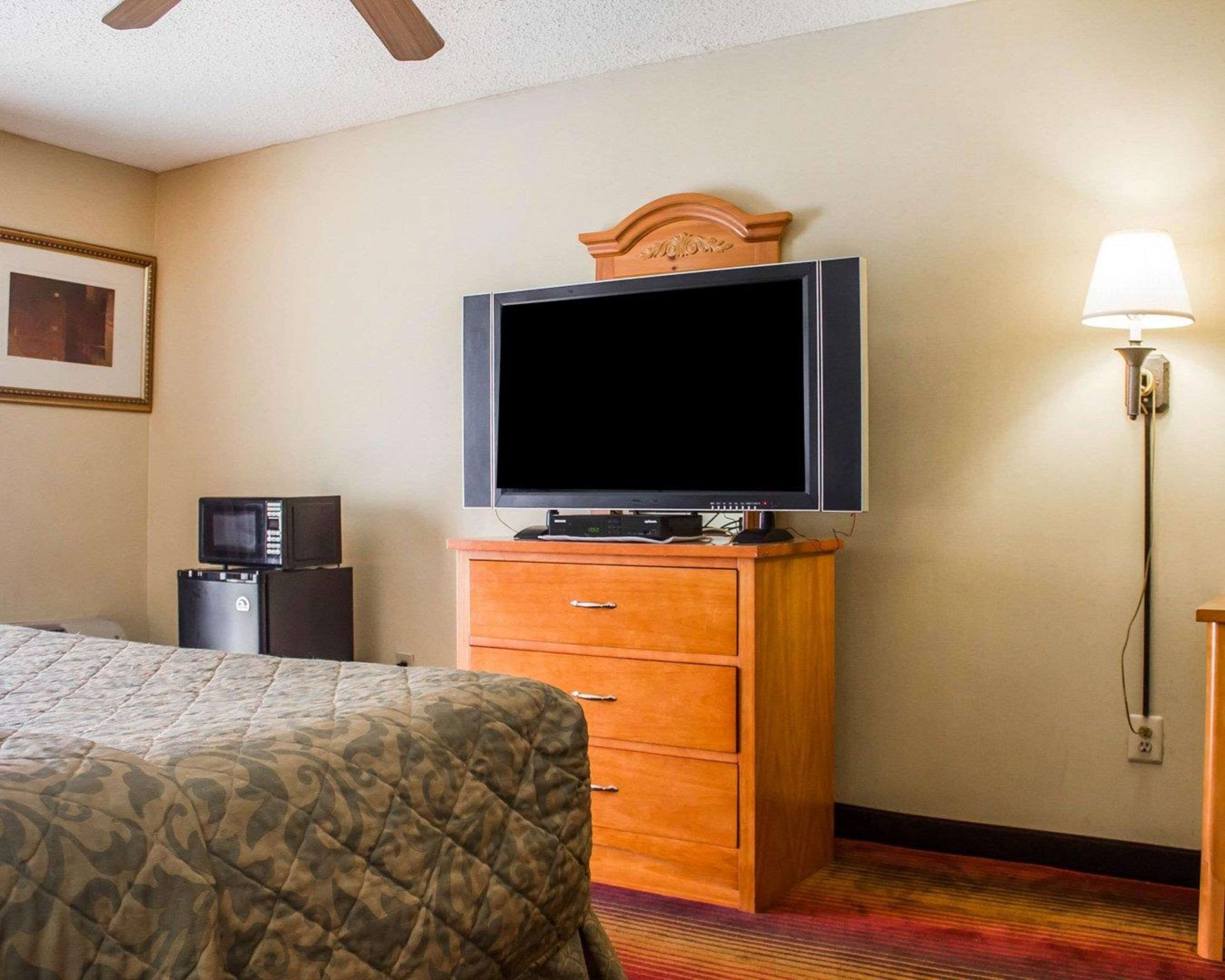 Quarto de hotel com cama, televisão sobre um suporte de madeira, micro-ondas, abajur e um quadro na parede.